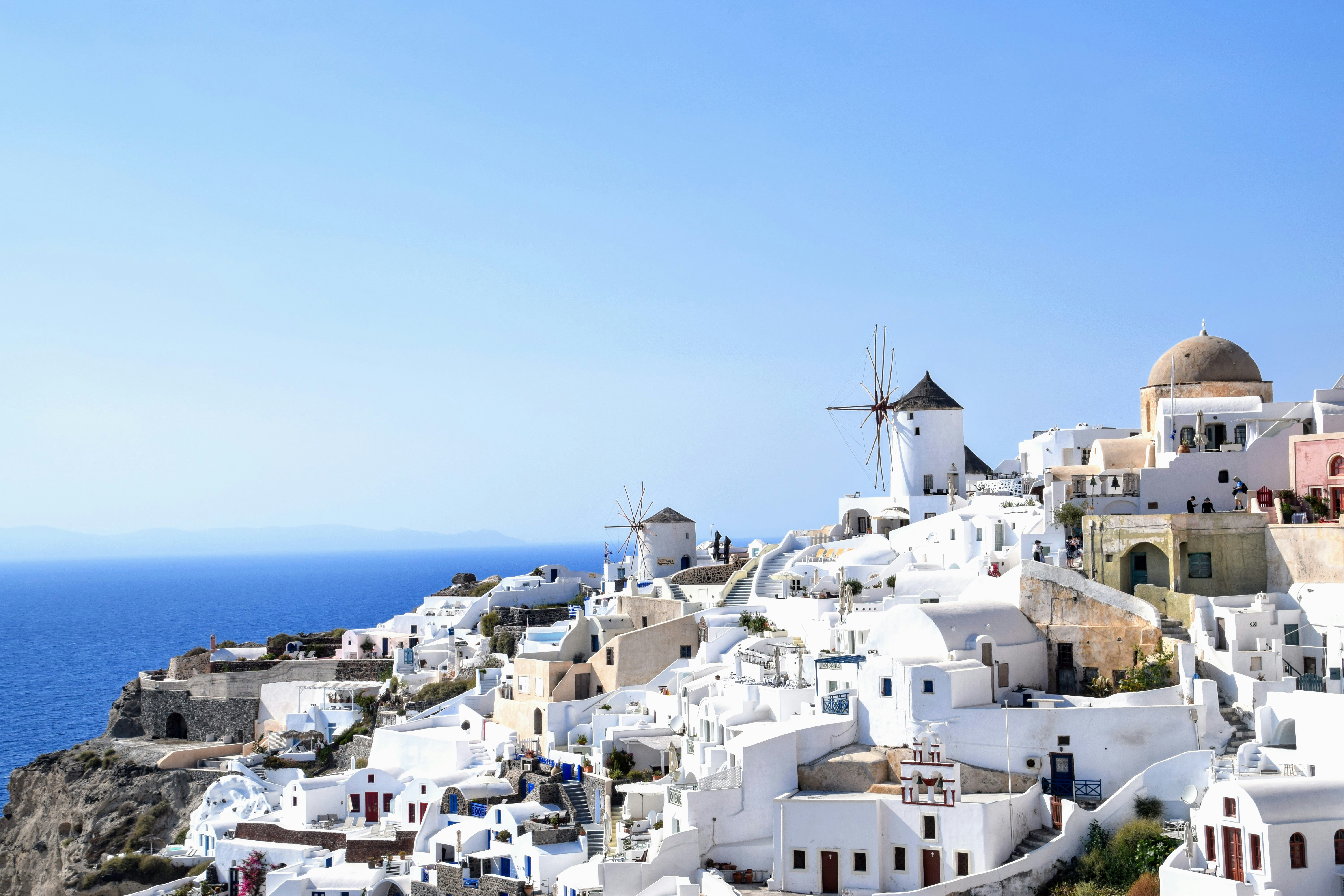 Whitewashed buildings and windmills overlook the deep blue Aegean Sea under a clear sky.