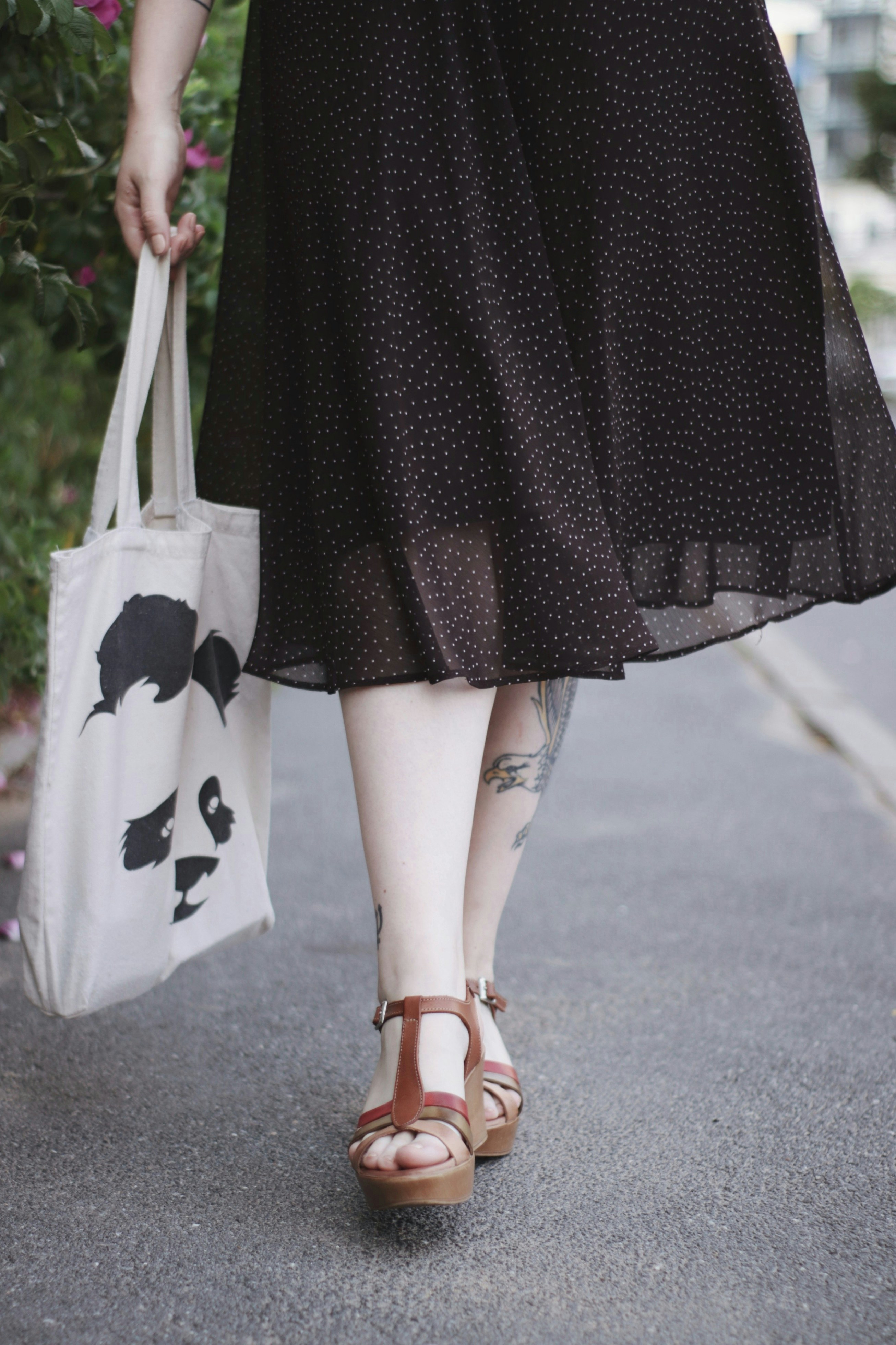 A woman walks down a city sidewalk, wearing a polka dot dress and carrying a tote bag adorned with a panda illustration.