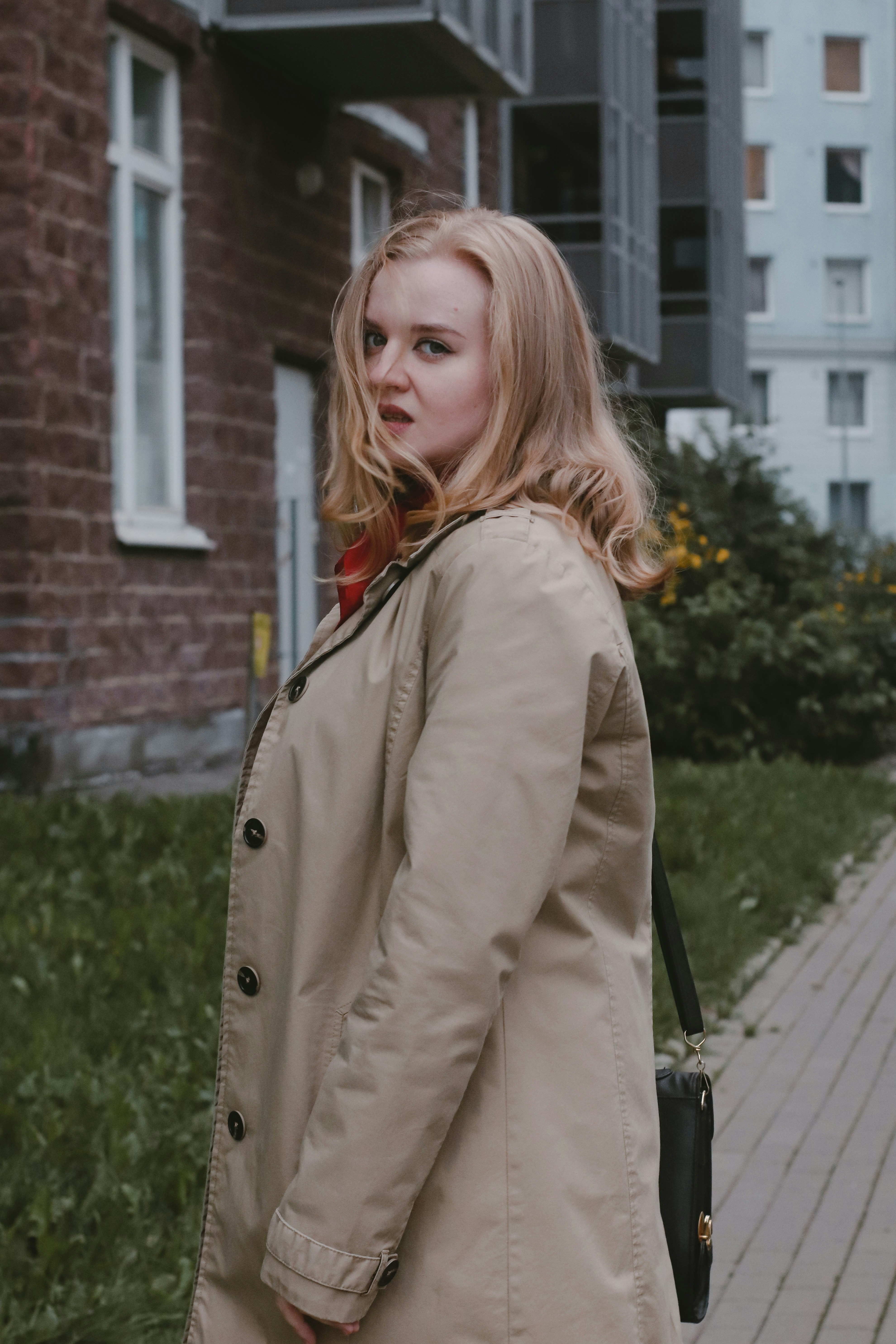Woman in a beige coat glances back while walking along a city pathway, framed by contemporary buildings and greenery.