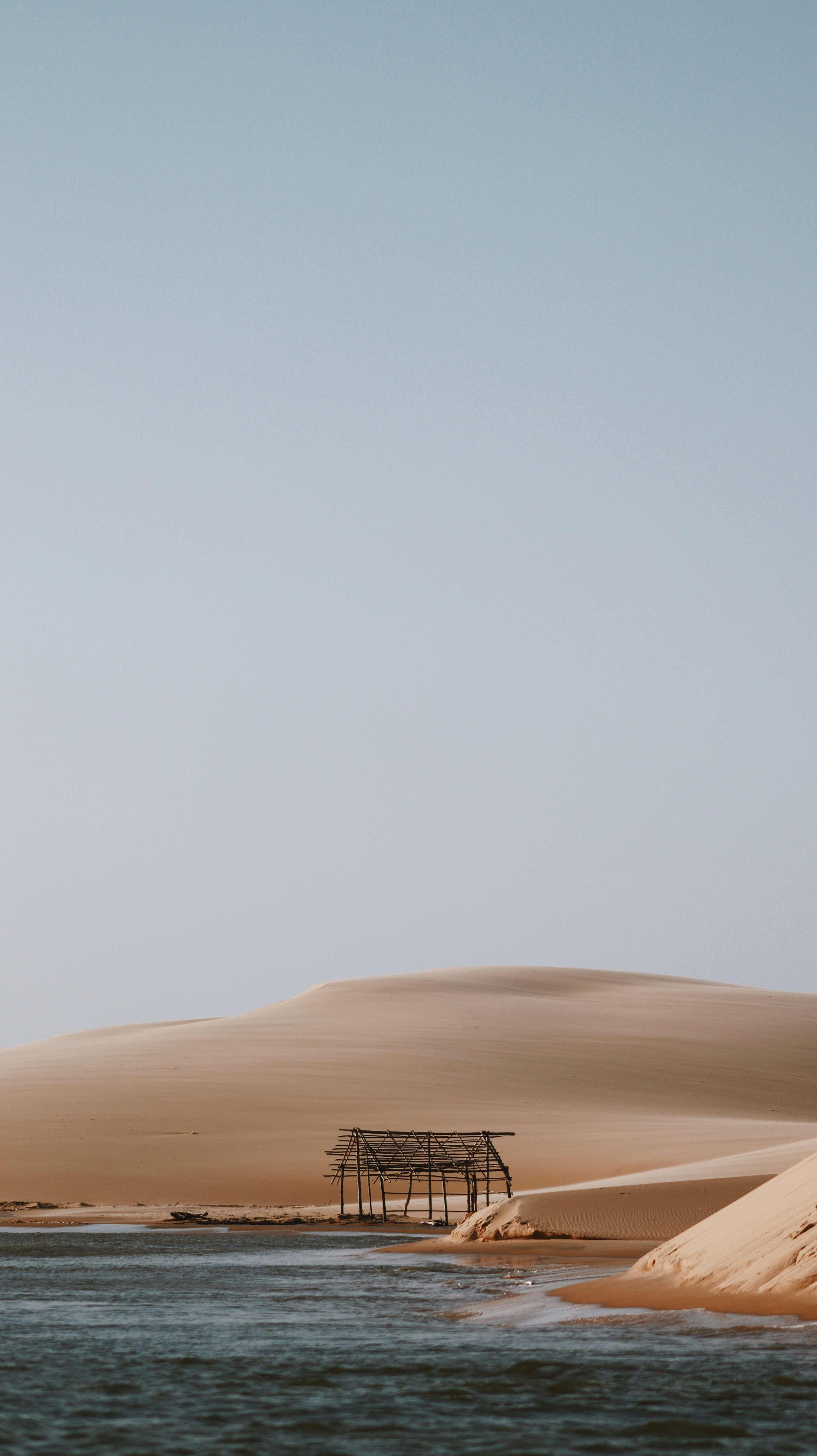 Abandoned wooden structure stands at the edge of a serene waterway, framed by sweeping sand dunes under a clear sky.