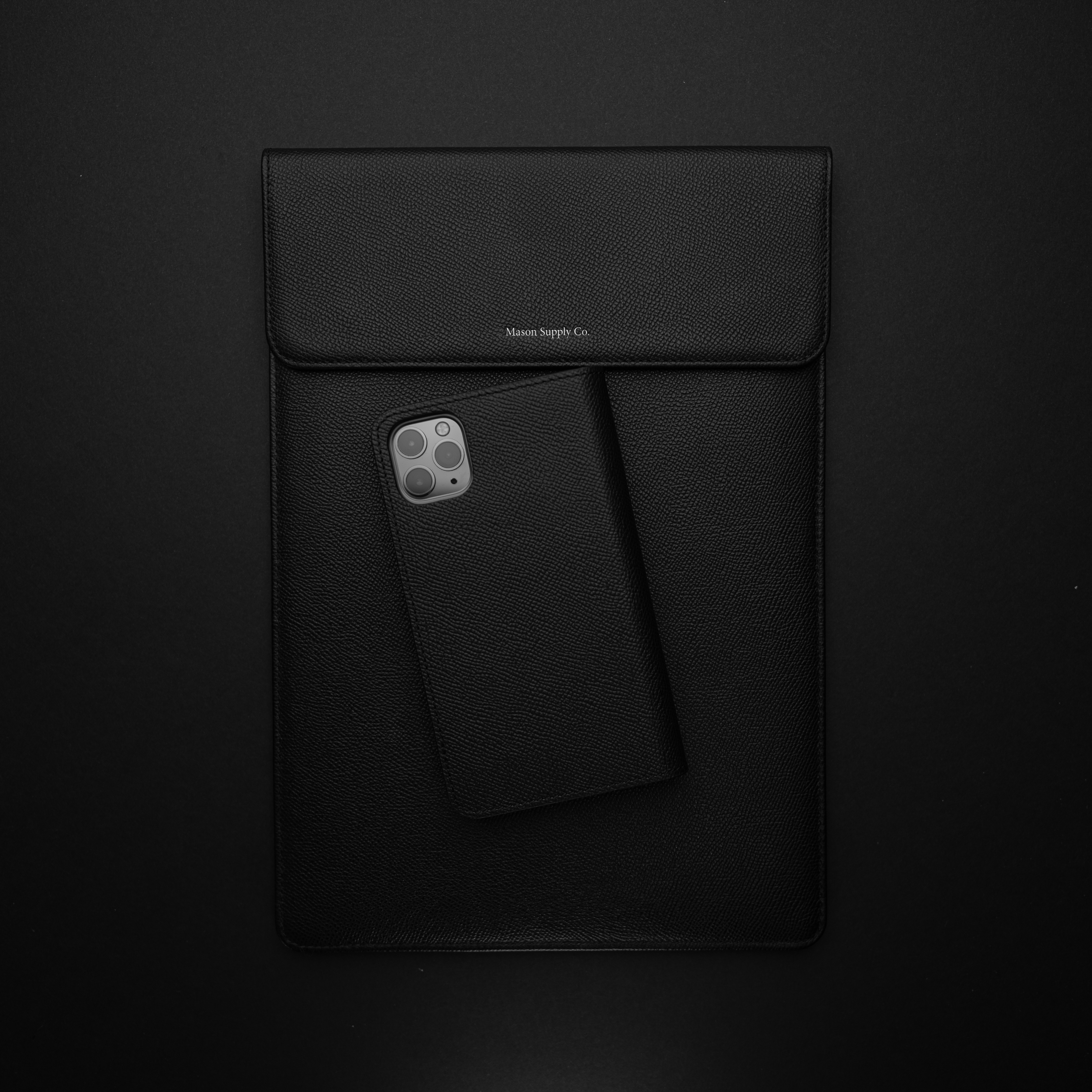 Monochrome product photograph of a black leather wallet with a diagonally placed phone case on top. The image emphasizes texture and minimalist design against a dark background.