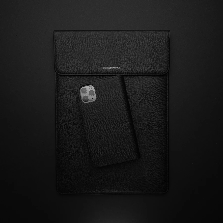A black leather folio and matching phone case arranged on a dark background, with a small white brand mark near the top flap. The composition is clean, monochrome, and understated.