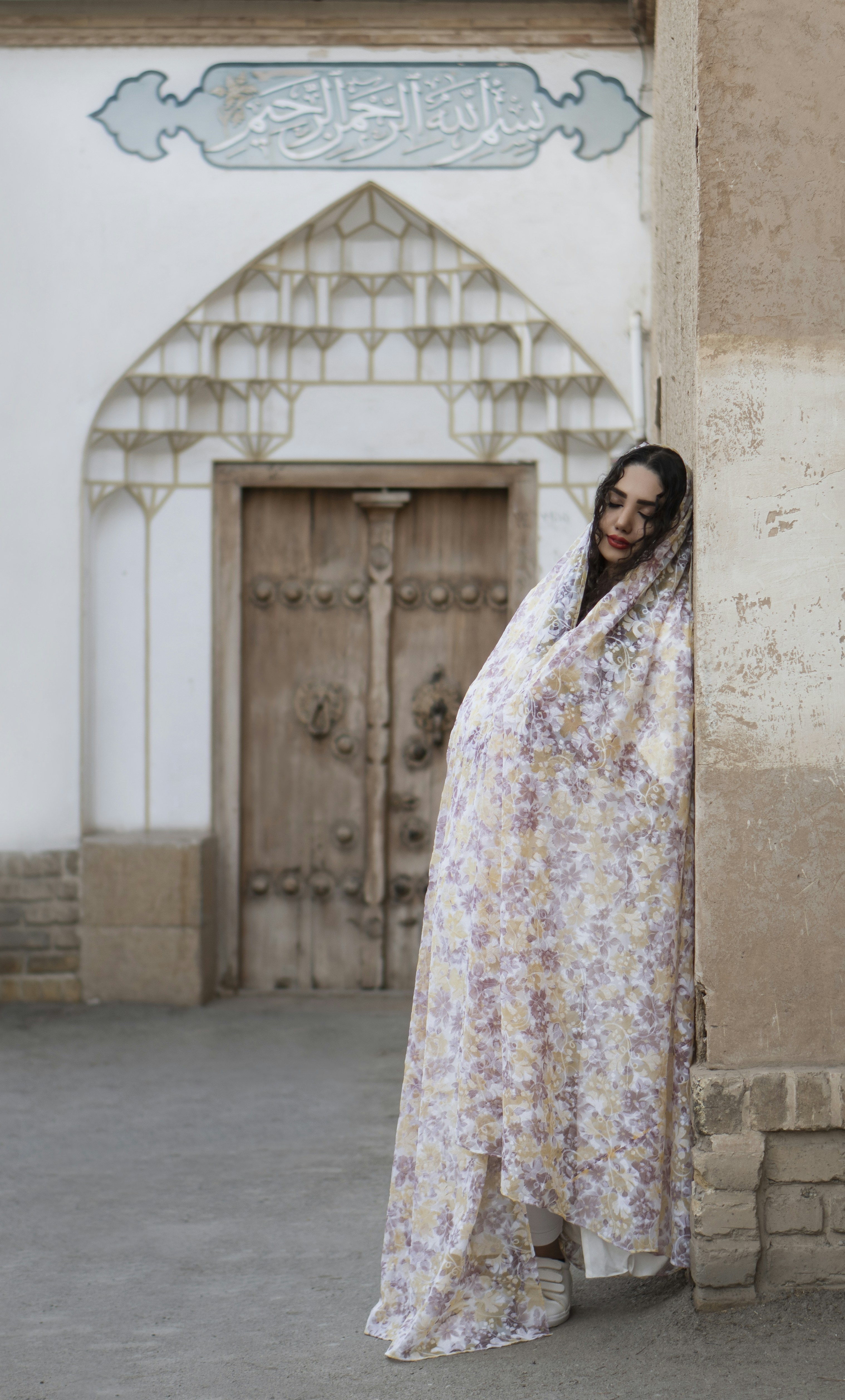 Woman in floral patterned hijab leaning against a rustic wall near an ornate wooden door.