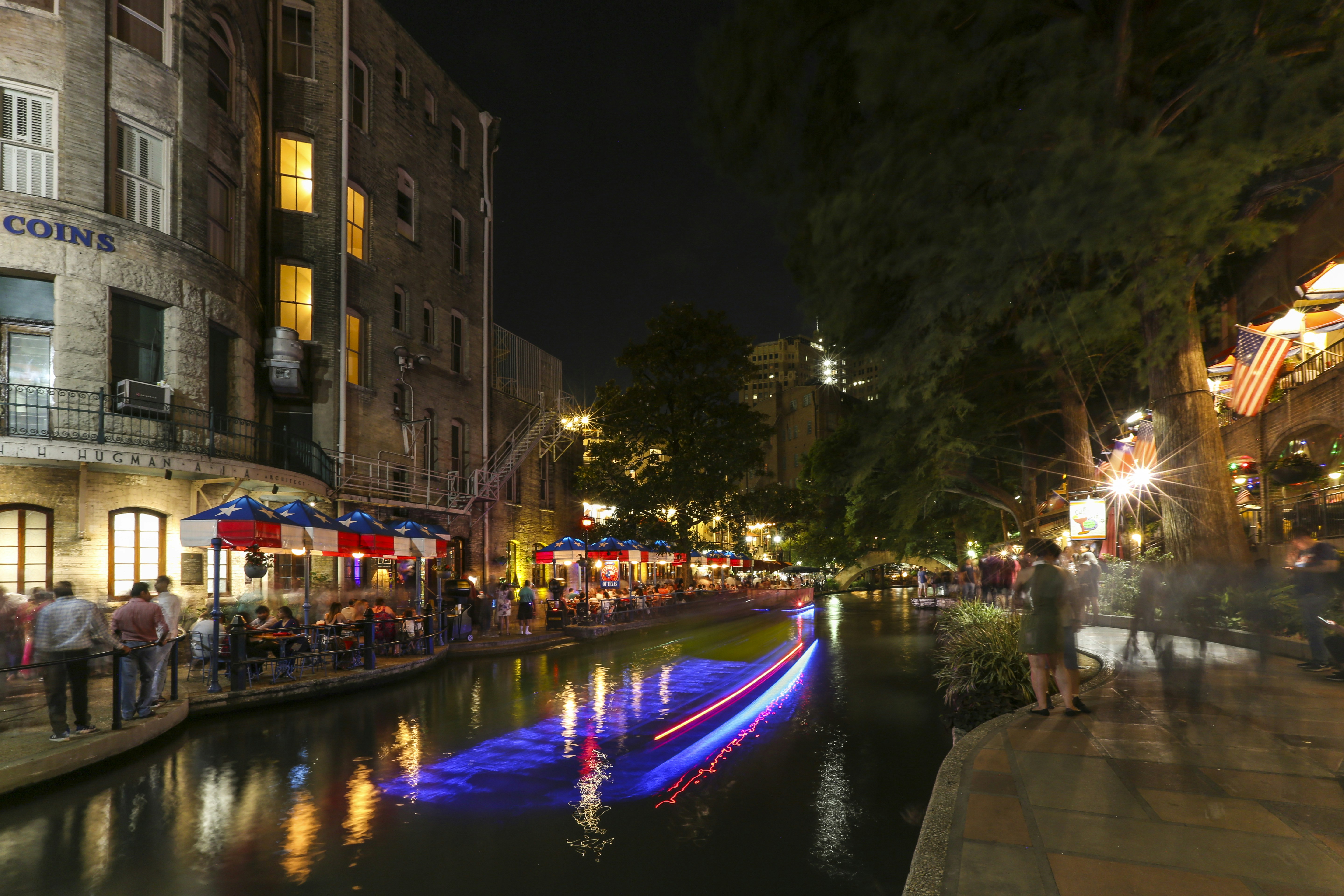People stroll along a riverside path illuminated by colorful lights from passing boats at night.