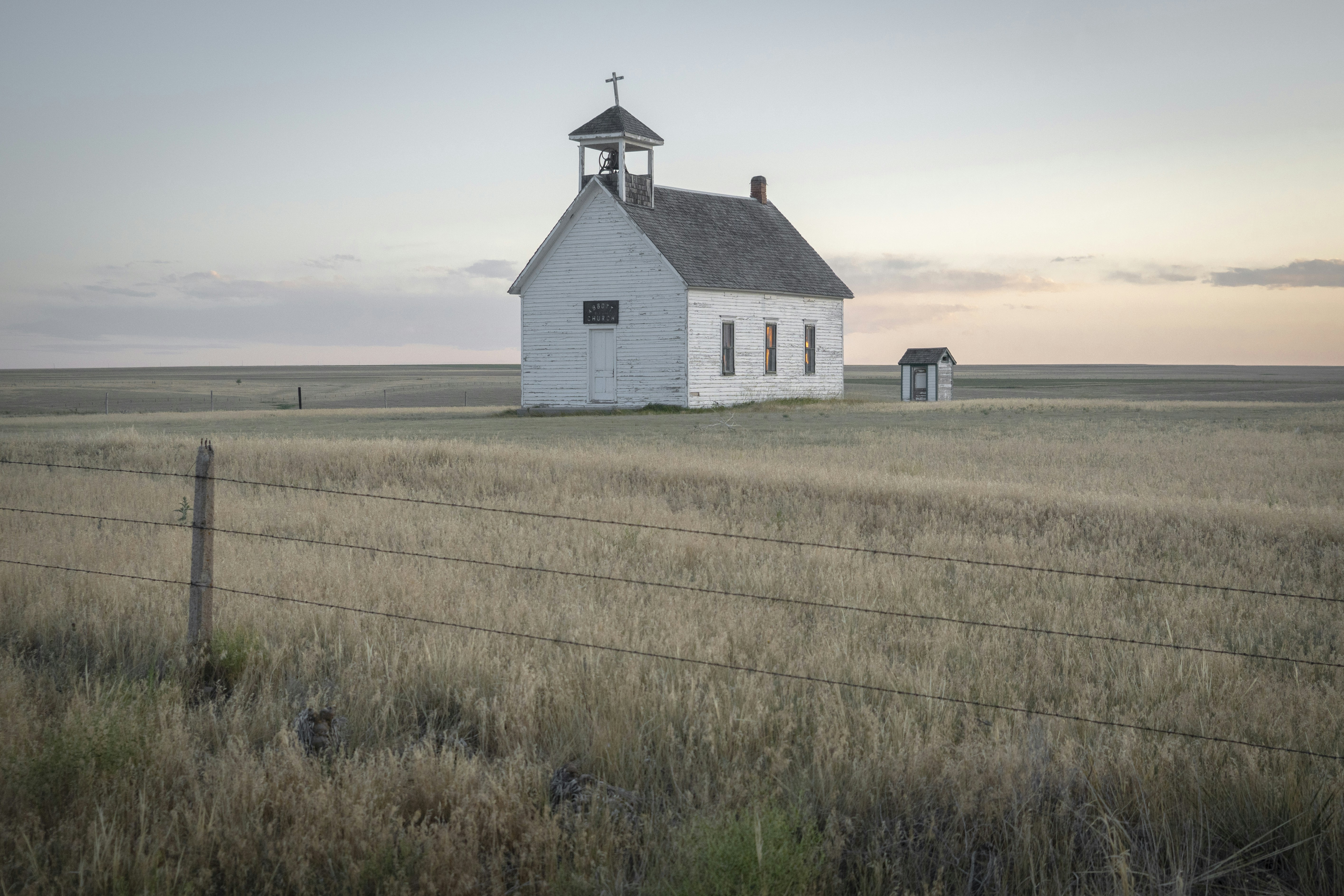 Weathered schoolhouse nestled in golden grasslands, evoking a sense of nostalgia and solitude. A small shed stands nearby under a soft, pastel sky.