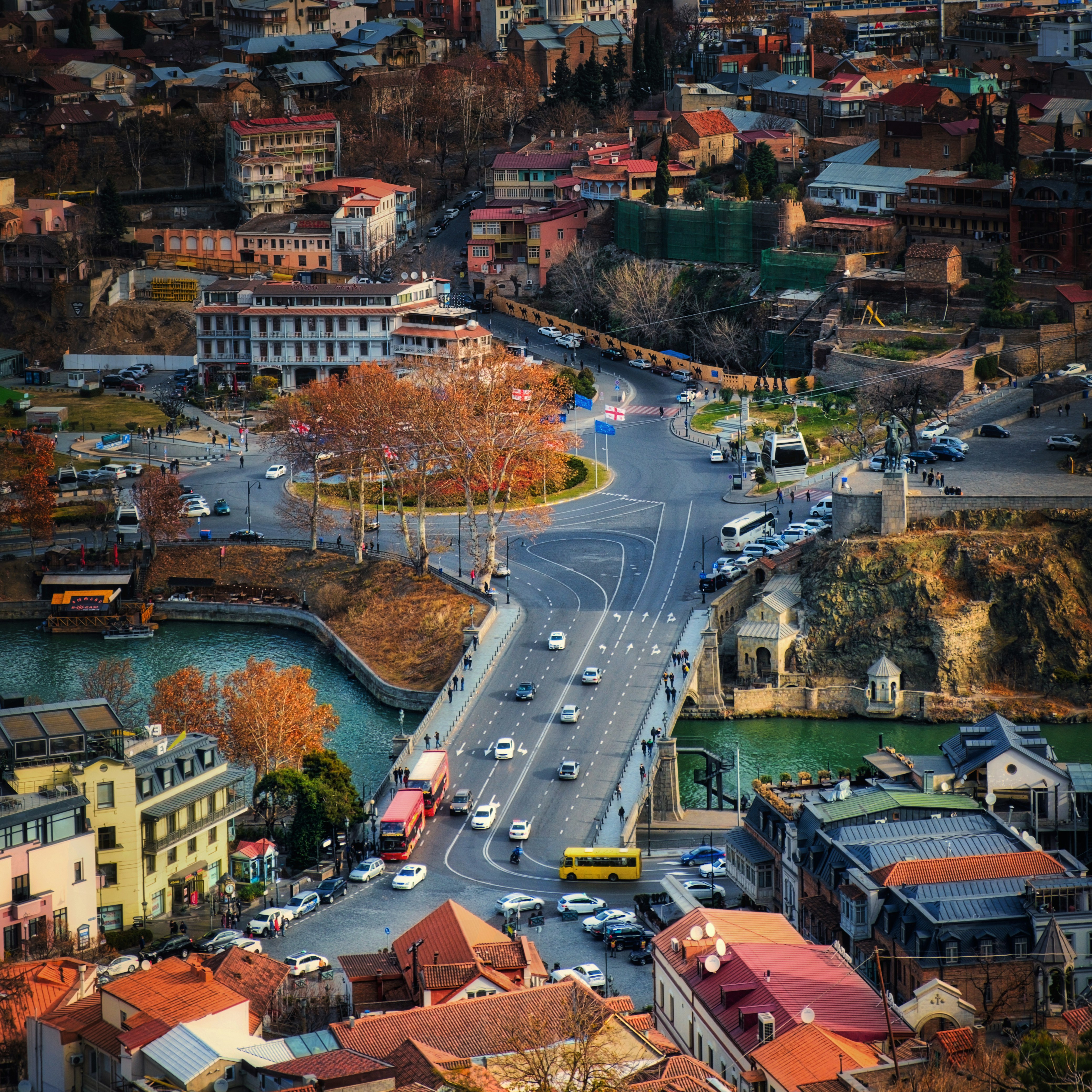 Aerial view of Tbilisi showcasing a busy intersection surrounded by colorful buildings and lush trees. The scene reflects the vibrant urban life and architecture of the city.