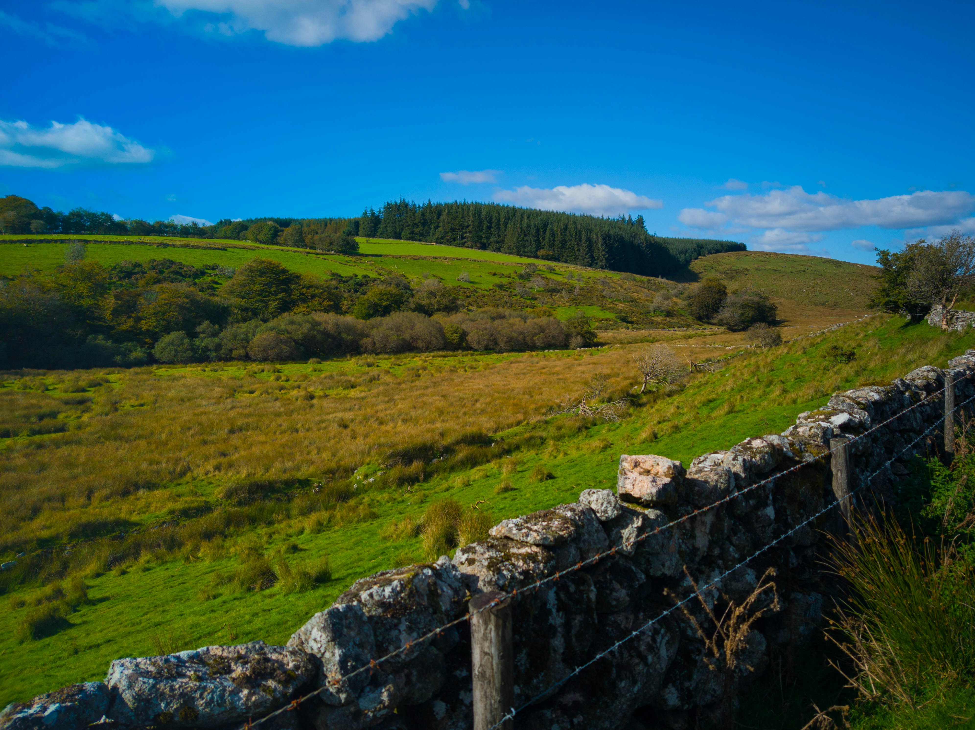 Lush green hills under a clear blue sky, framed by a rustic stone fence, create a peaceful rural landscape.