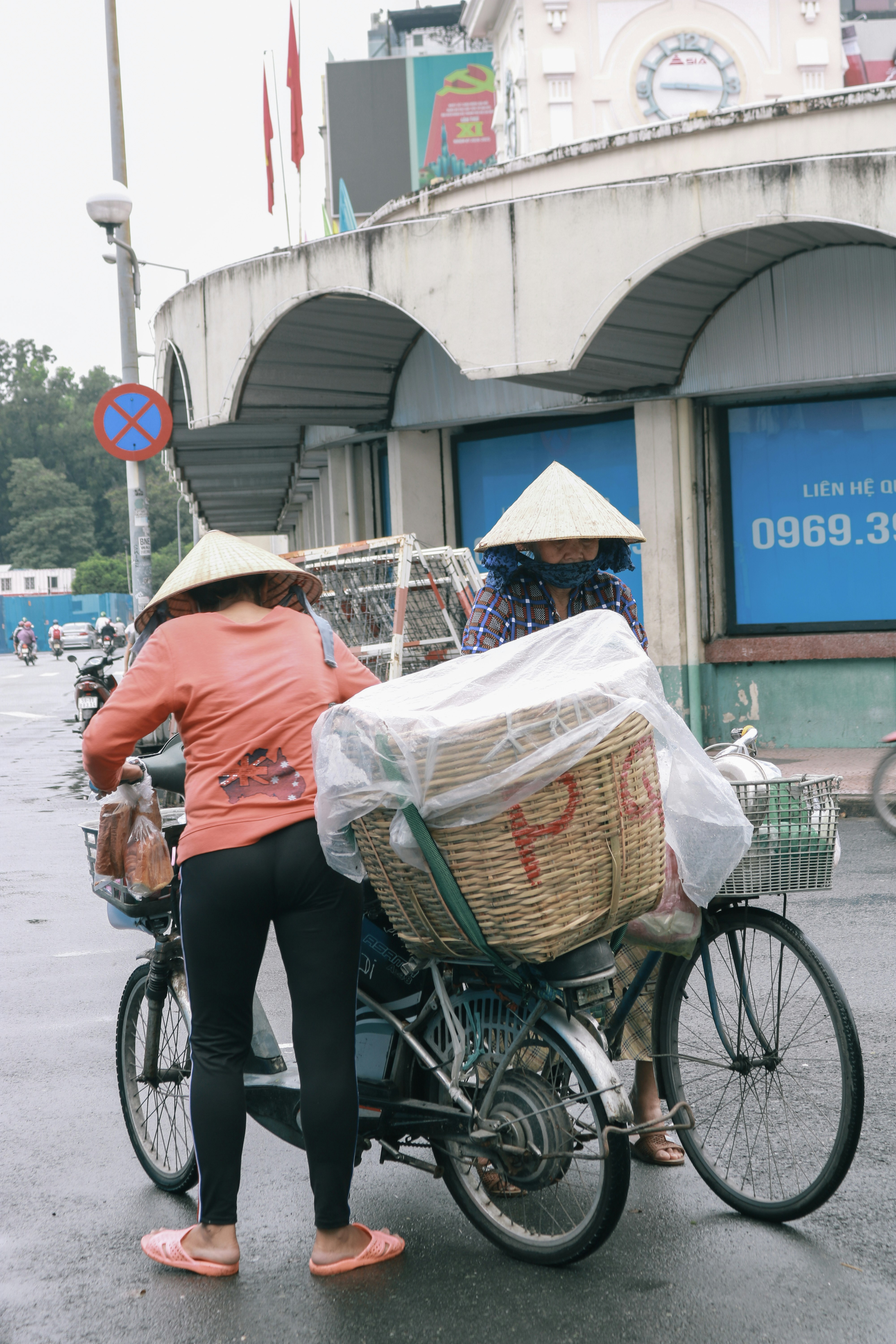 Two women in conical hats adjusting a bicycle laden with woven baskets in a bustling urban environment.