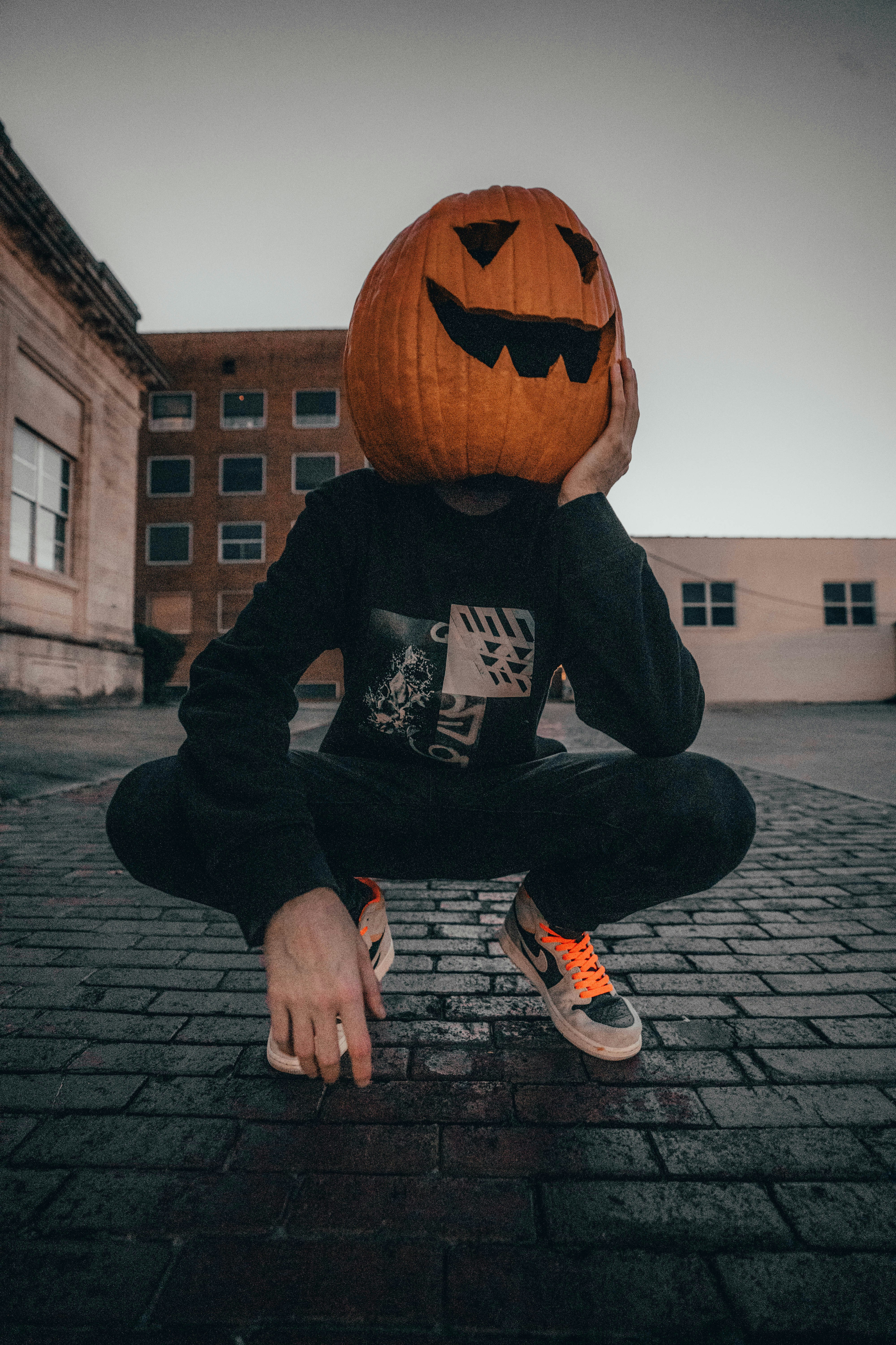 Figure squatting with a carved pumpkin on their head, set against an urban backdrop at dusk.