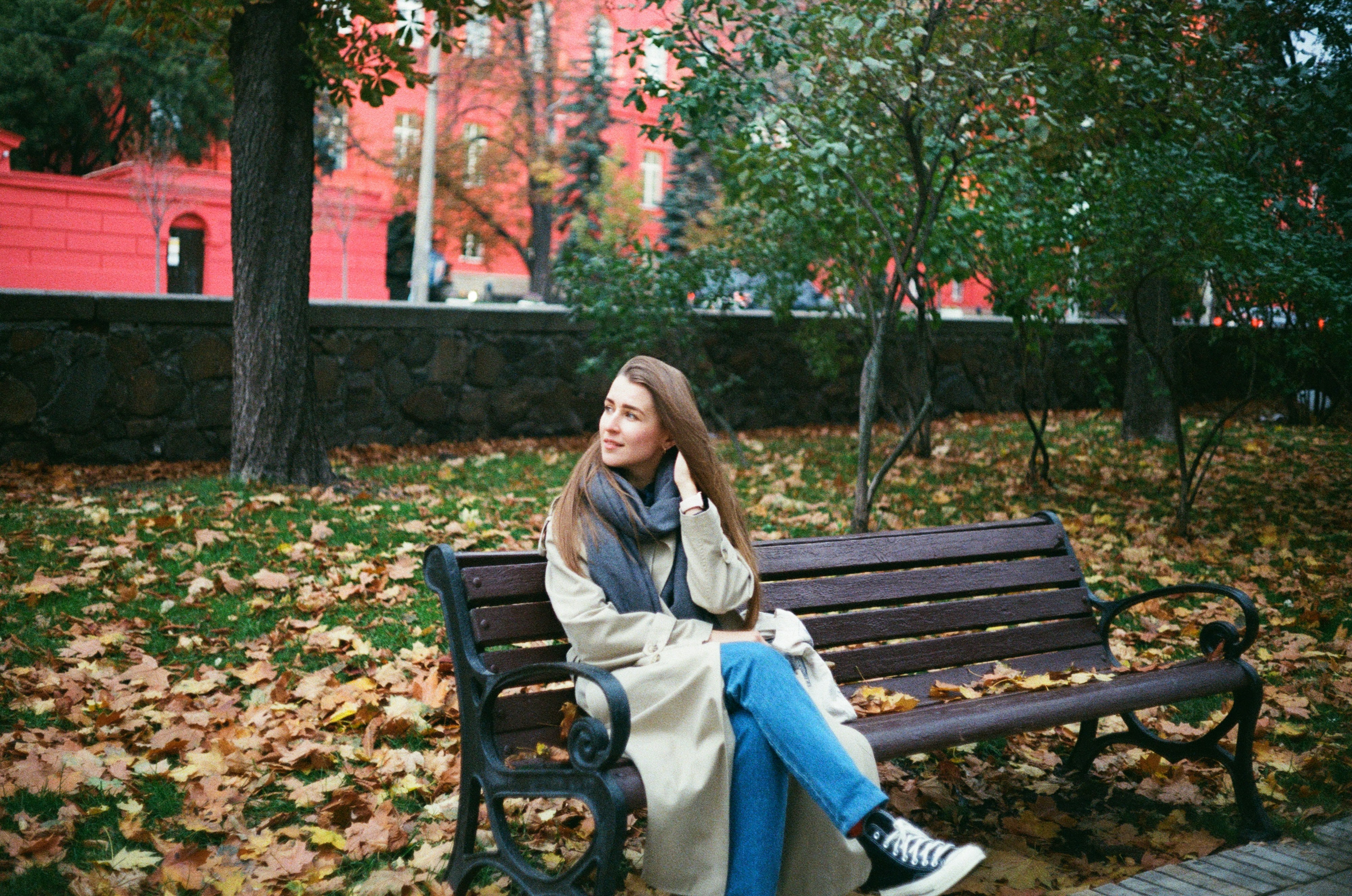 A candid photograph of a woman seated on a park bench amid autumn leaves, wearing a beige coat and blue jeans, with a red brick backdrop.