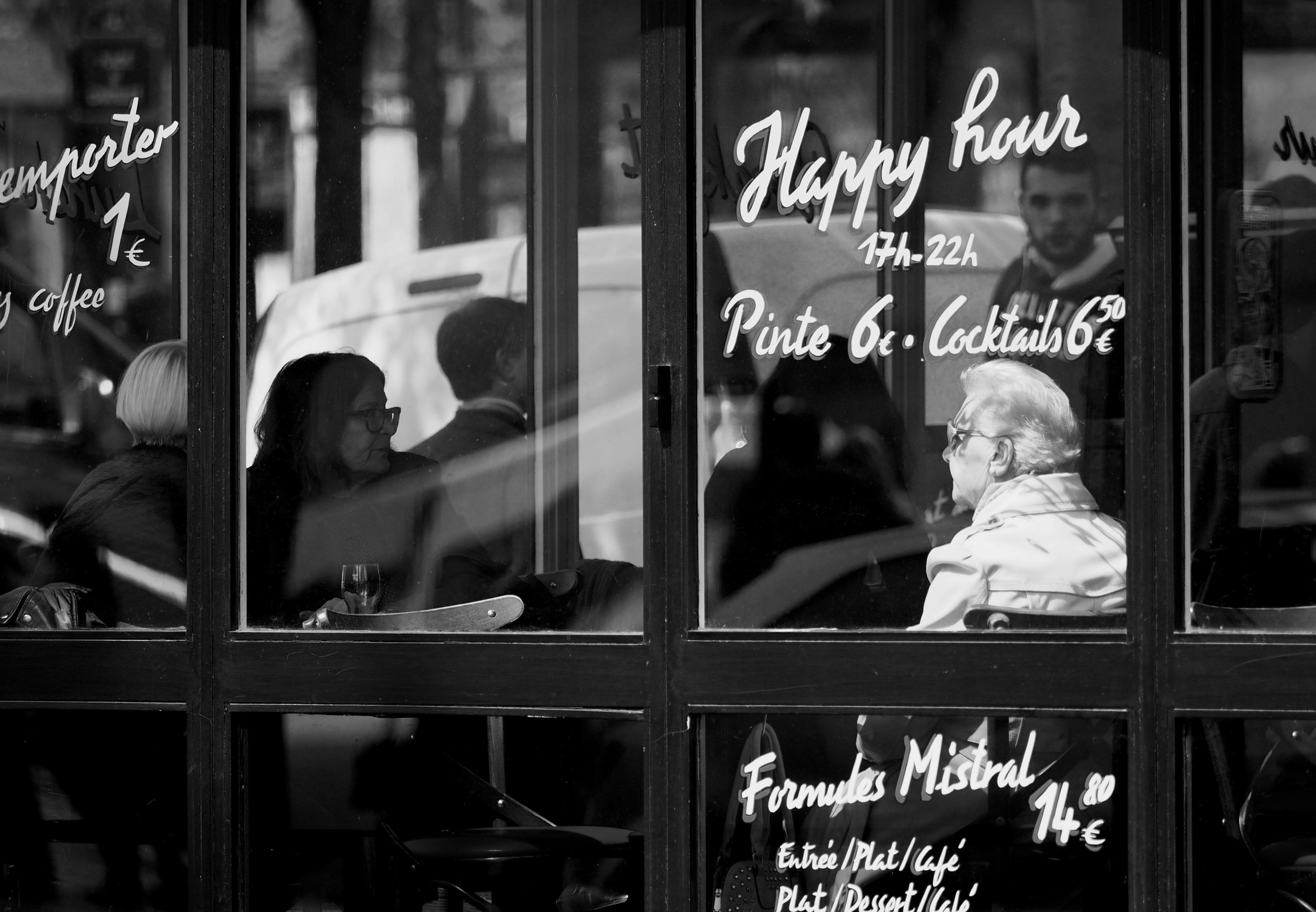 Black and white scene of people conversing inside a café, framed by glass windows with handwritten menu details.