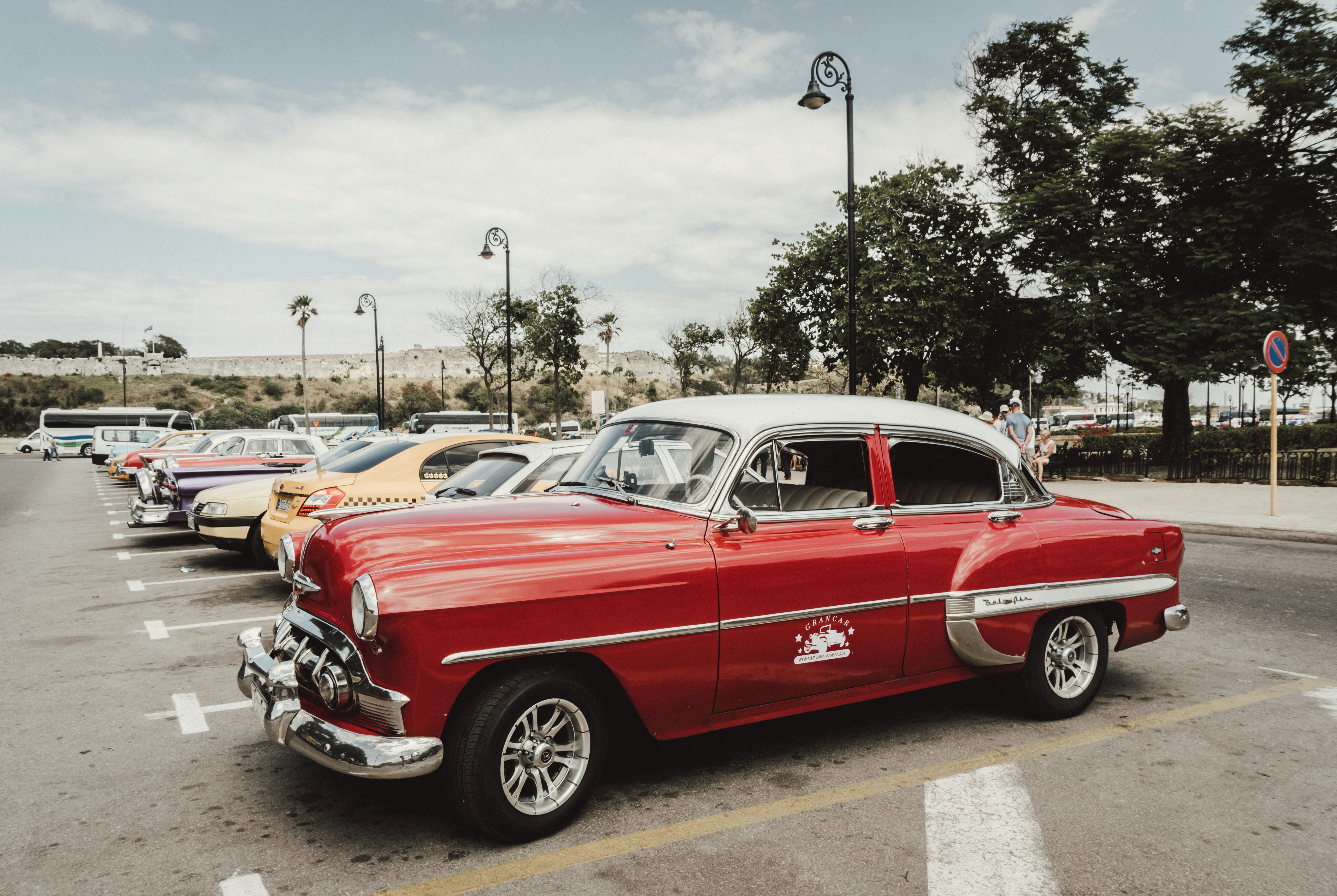 This captivating image showcases a row of vintage cars, dominated by a striking red classic, set against a serene backdrop of blue skies and lush greenery. The vibrant colors of the vehicles contrast beautifully with the muted tones of the surrounding environment, creating a nostalgic and lively atmosphere. The composition draws the eye along the line of cars, while the soft, natural lighting highlights the glossy paintwork and chrome details, making the scene visually arresting.