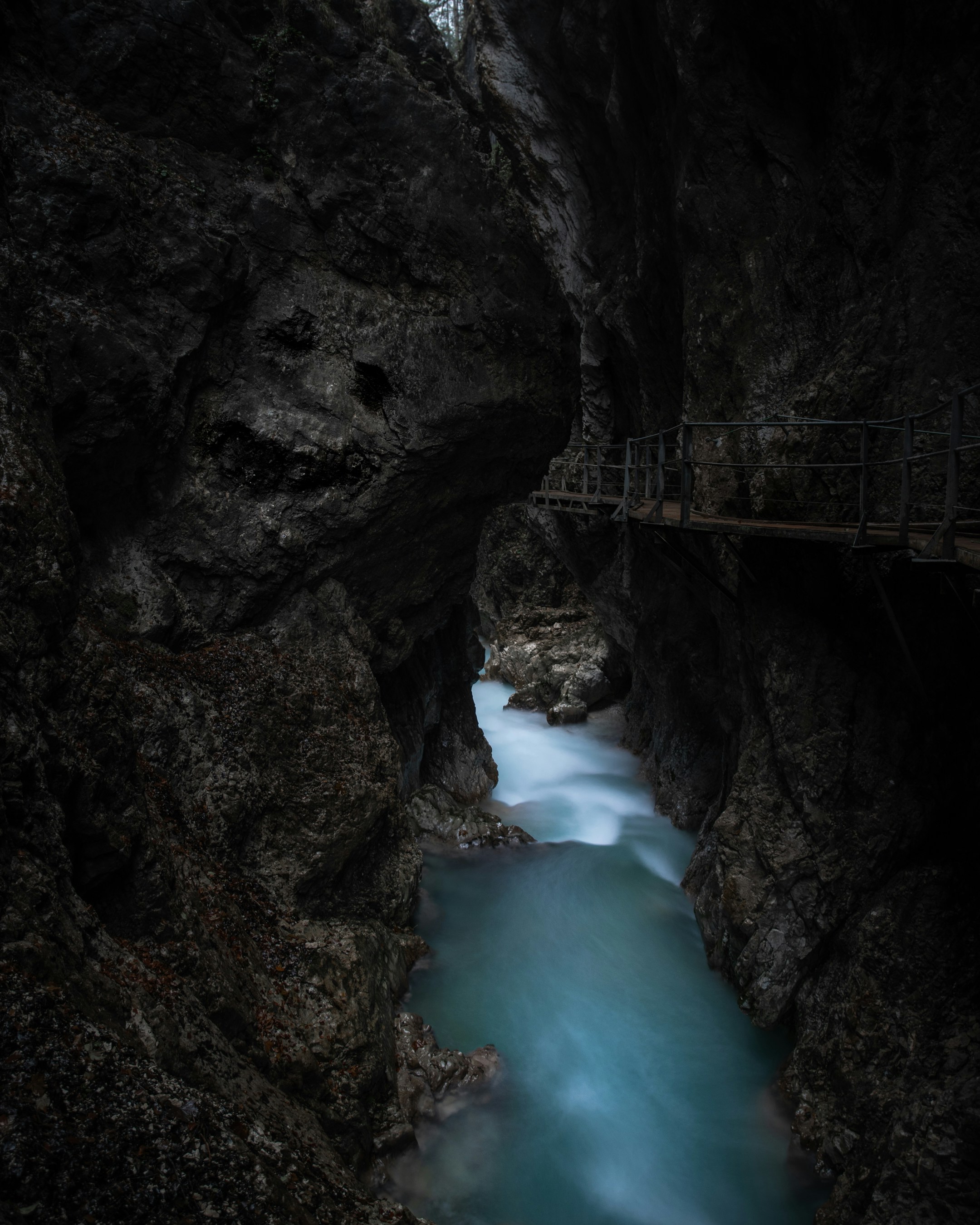 Long-exposure photograph of a turquoise river winding through a dark rock canyon, with a wooden walkway clinging to the right wall.