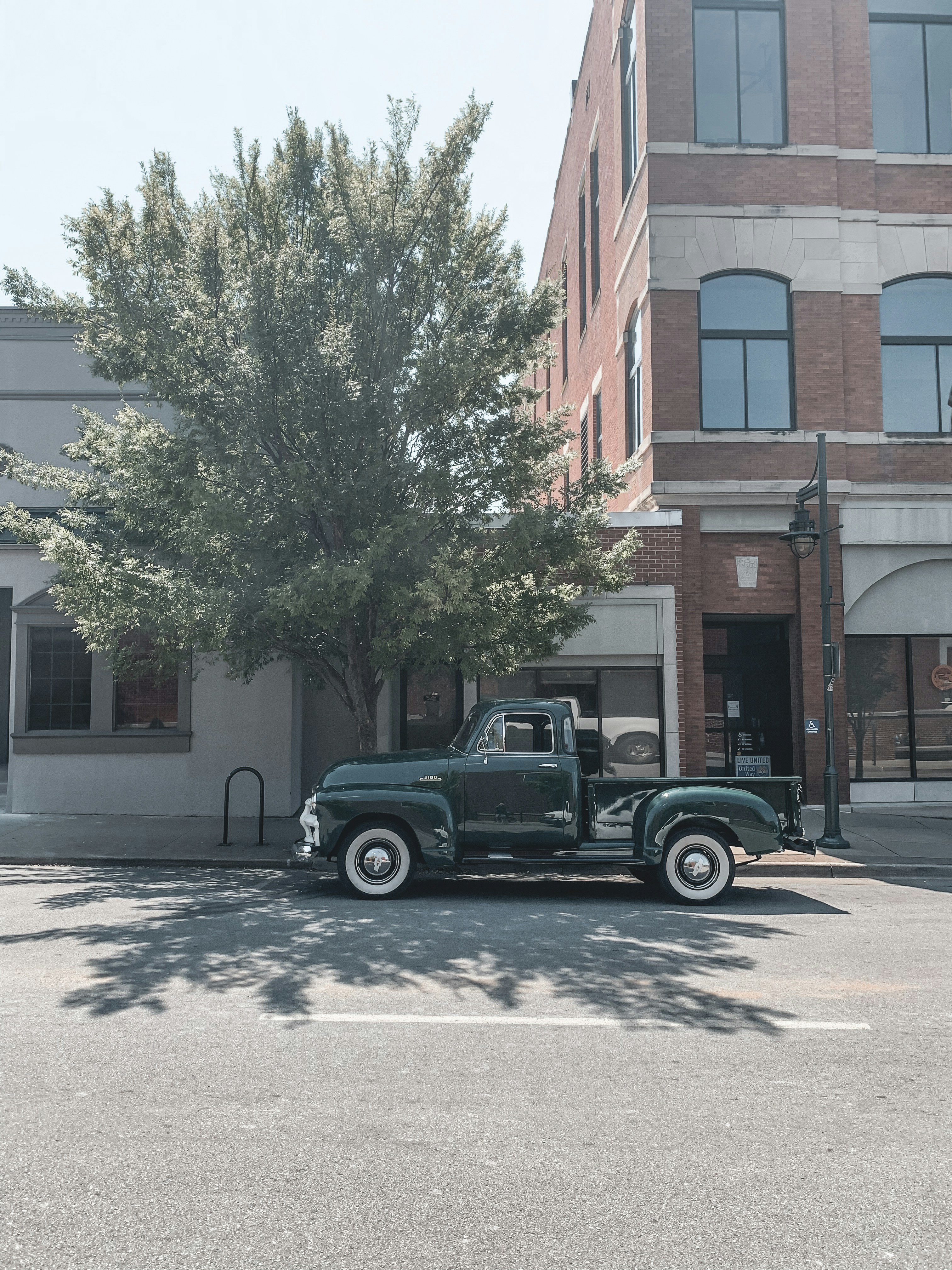 Vintage green pickup truck parked under a leafy tree beside a historic brick building.