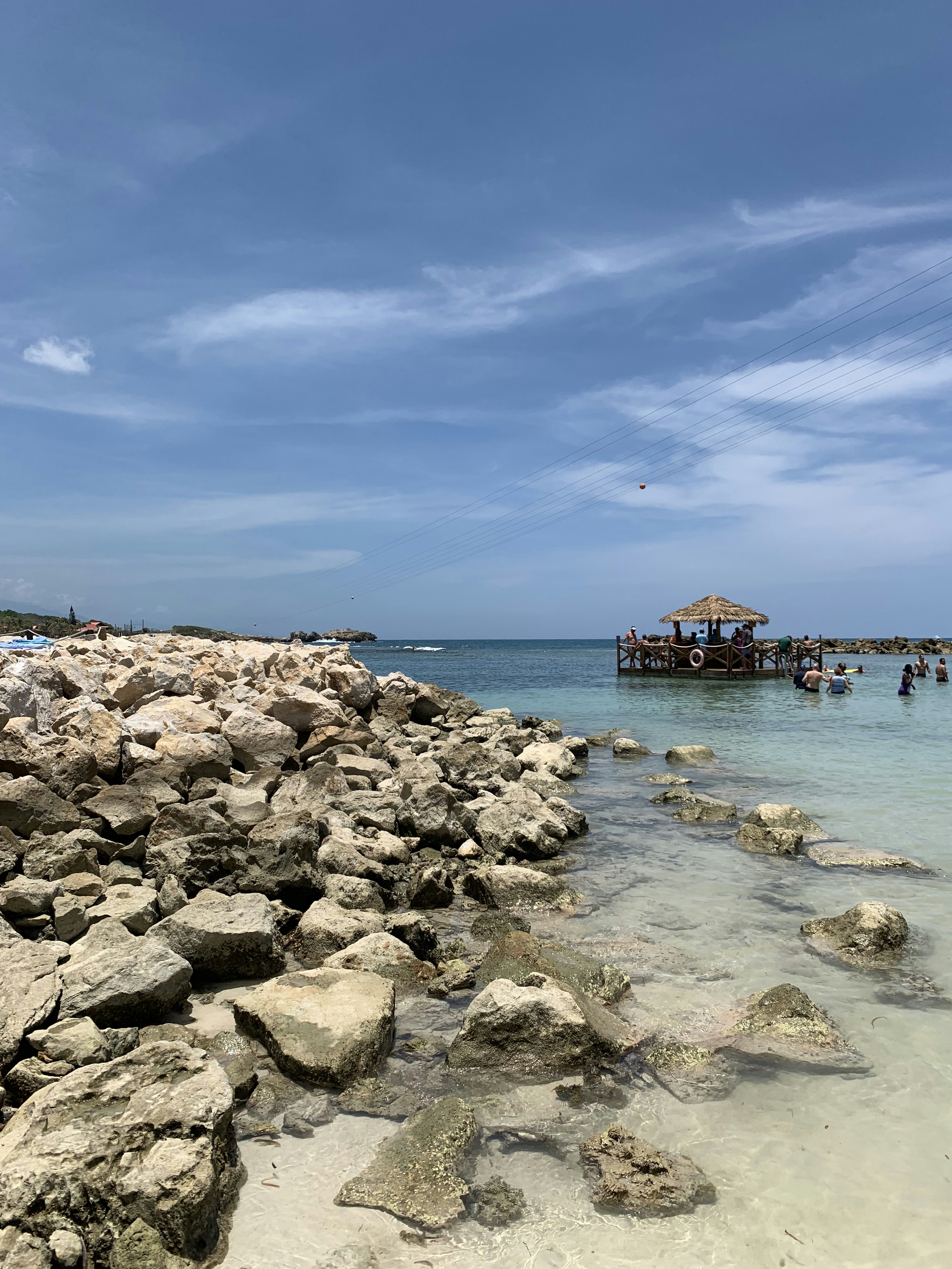 Rocky shoreline leading to a dock with people enjoying the clear waters under a blue sky.