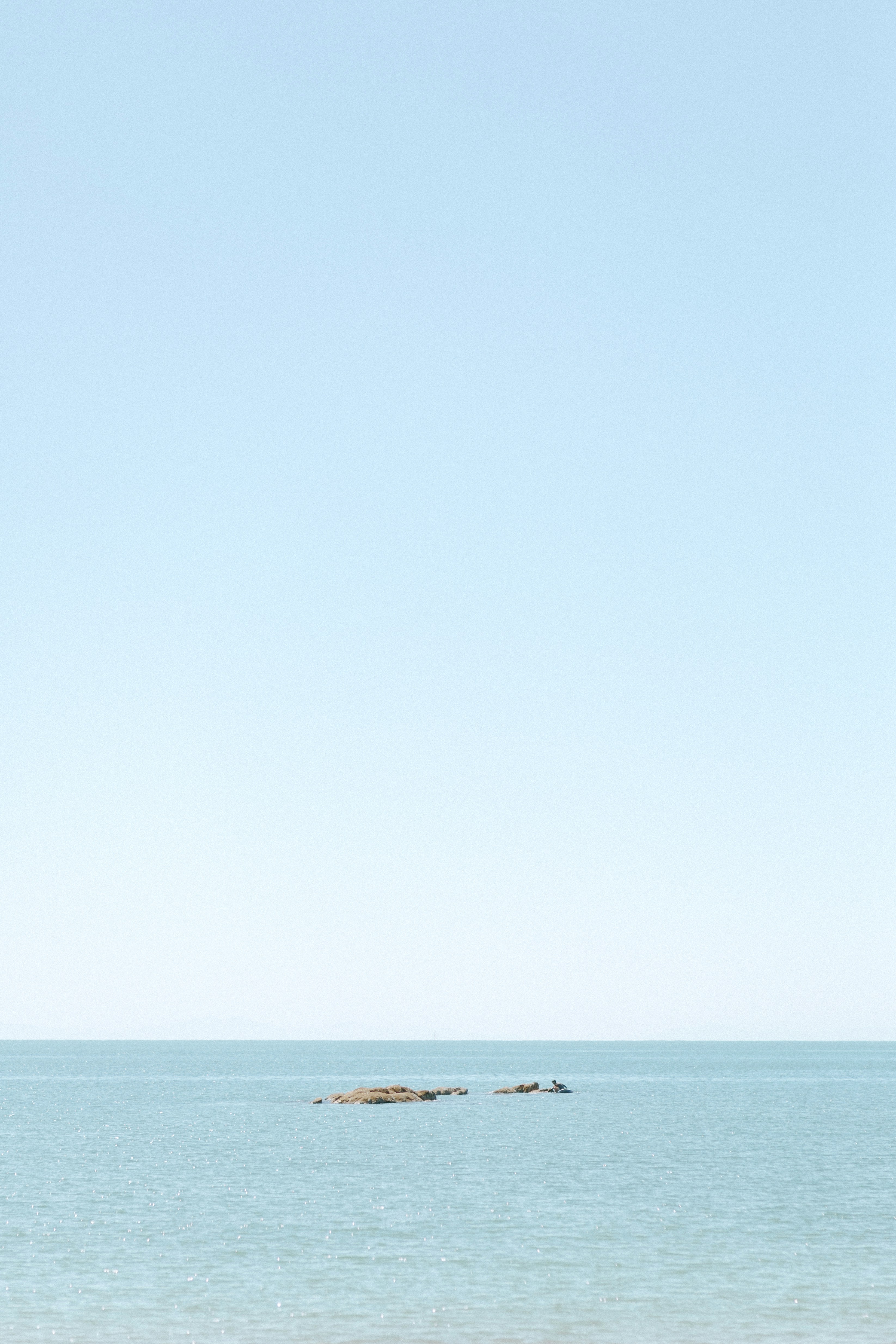 Calm sea meeting a clear sky, with distant rocks barely breaking the surface. A tranquil scene evoking peace.