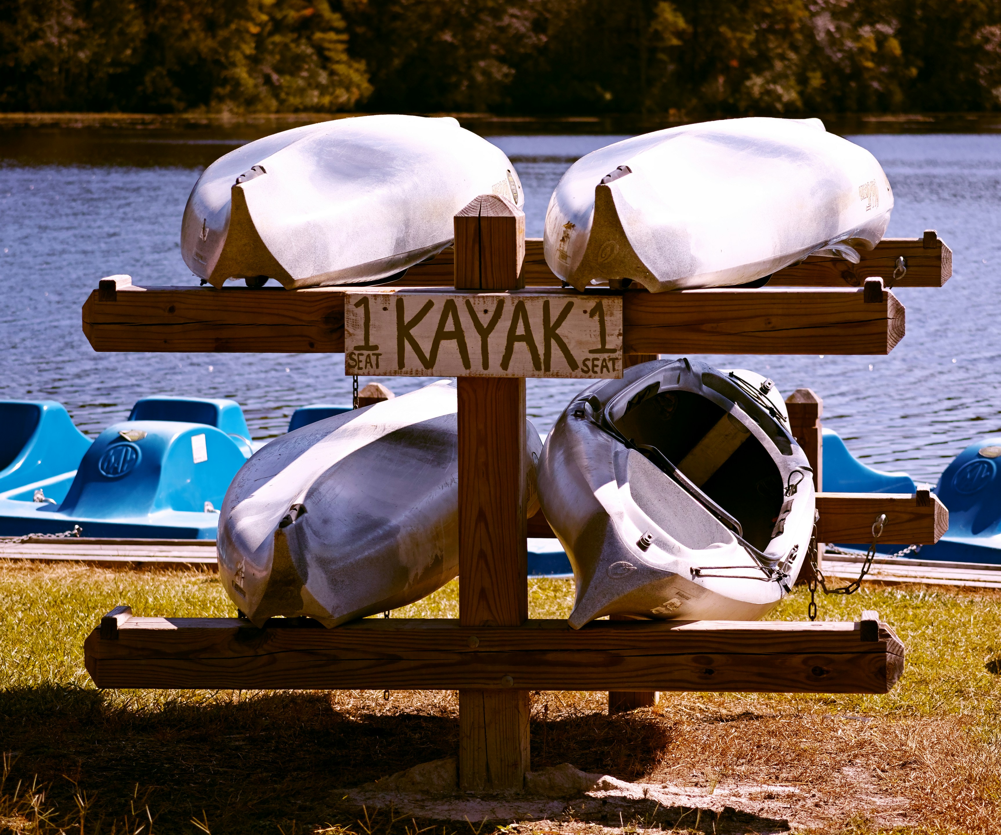 White kayaks stacked on a wooden rack by a lake, with blue pedal boats and calm water in the background.