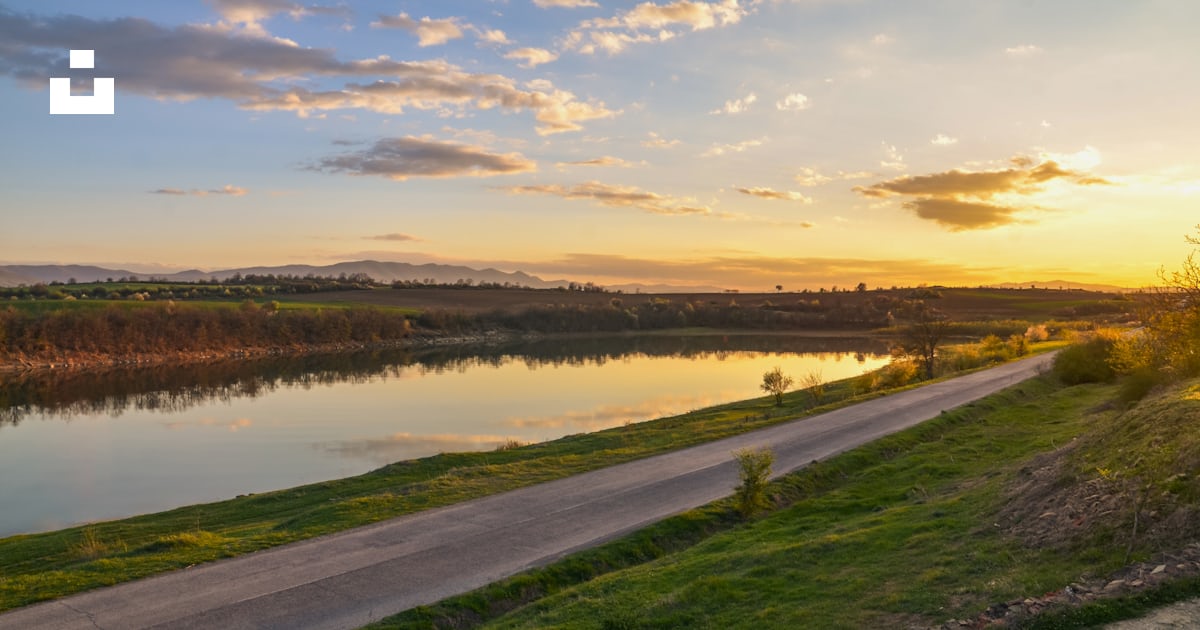 Green Grass Field Near Body Of Water During Sunset Photo Free Road