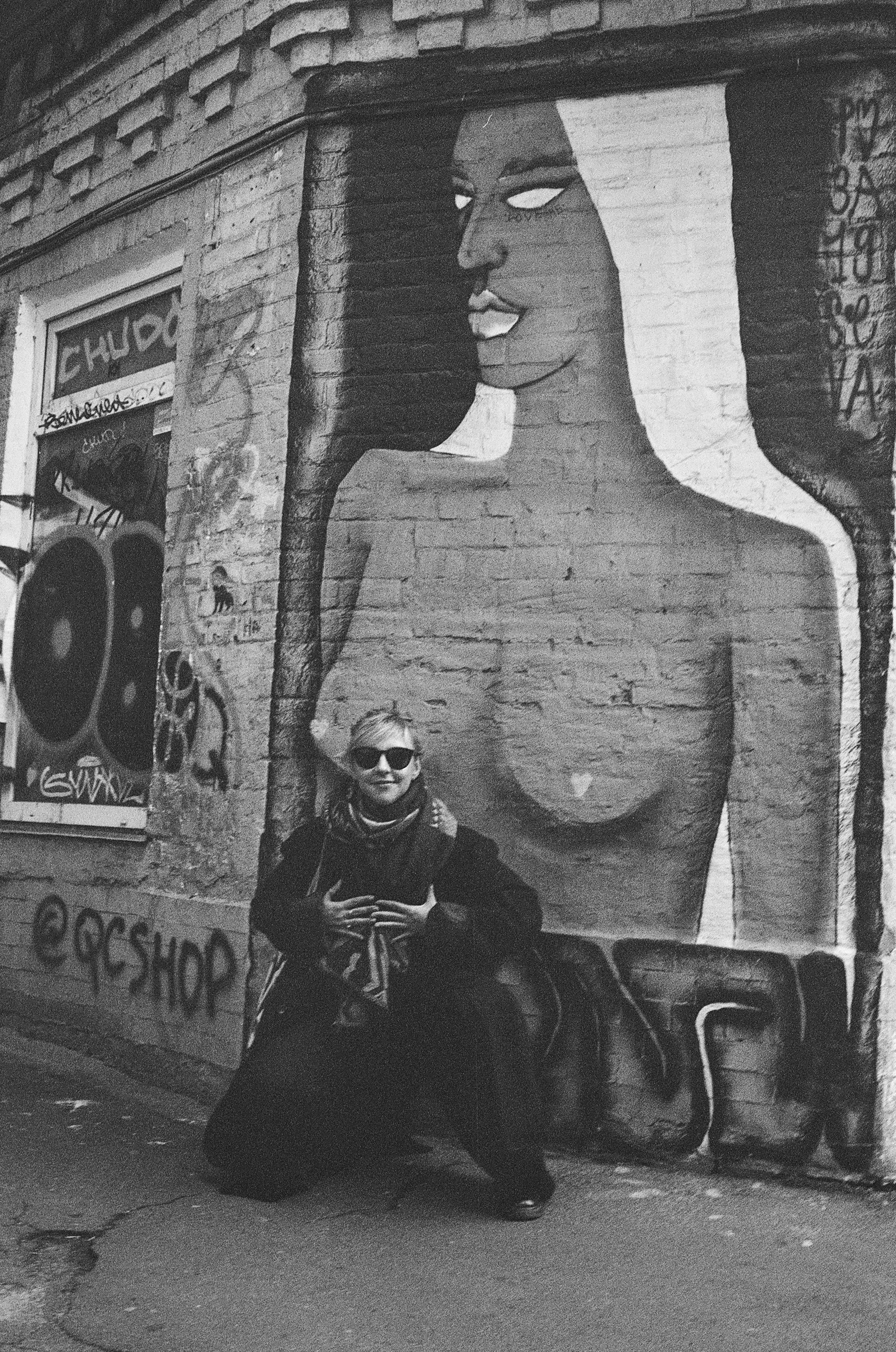 Black-and-white street photograph of a graffiti-covered wall with a large mural of a nude figure, and a person kneeling in front wearing sunglasses. The composition contrasts street art with a quiet human presence.