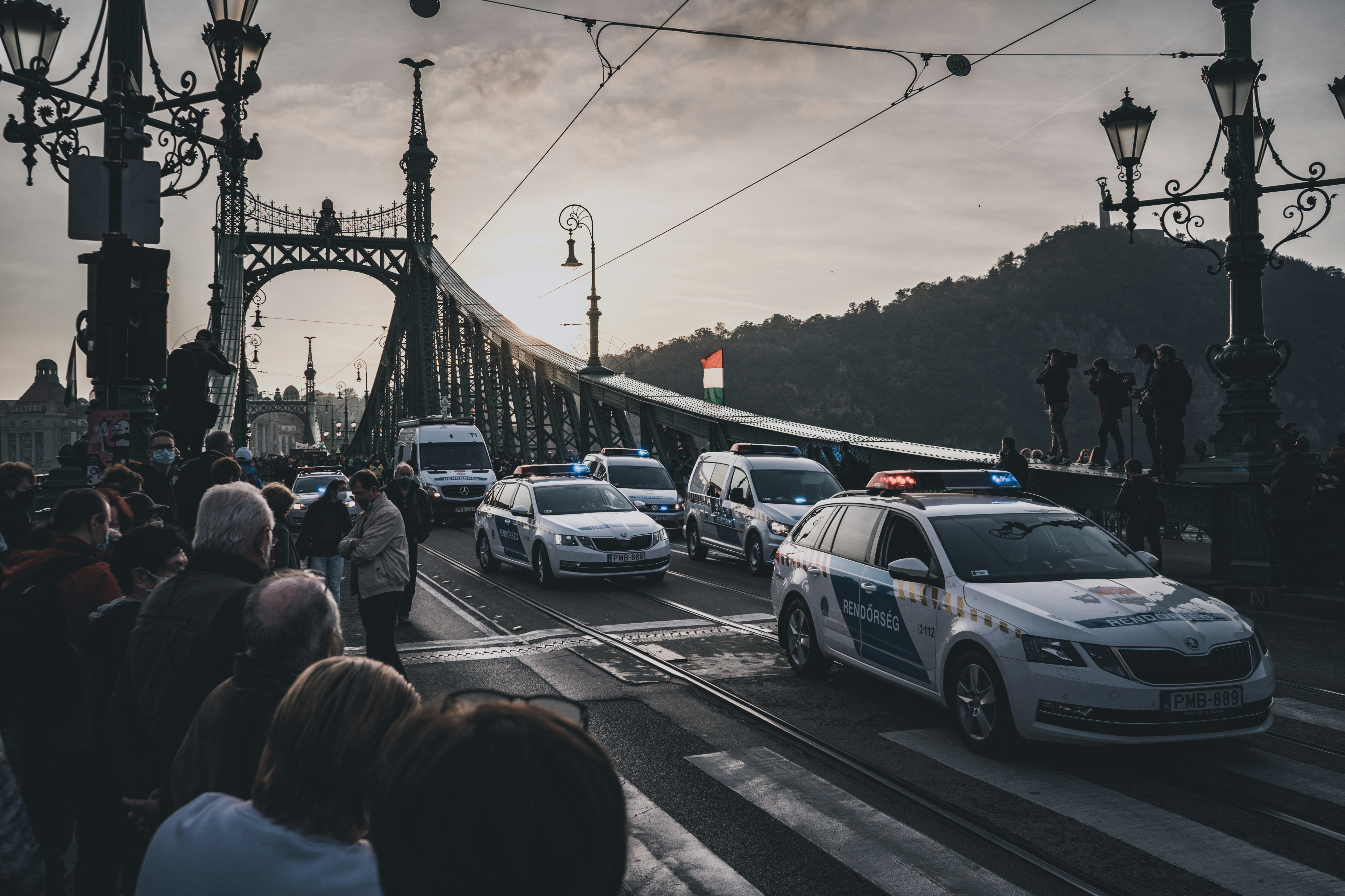 Police vehicles navigate a historic bridge while onlookers gather, capturing a moment of urban life and security. The scene reflects a blend of architecture and community engagement.