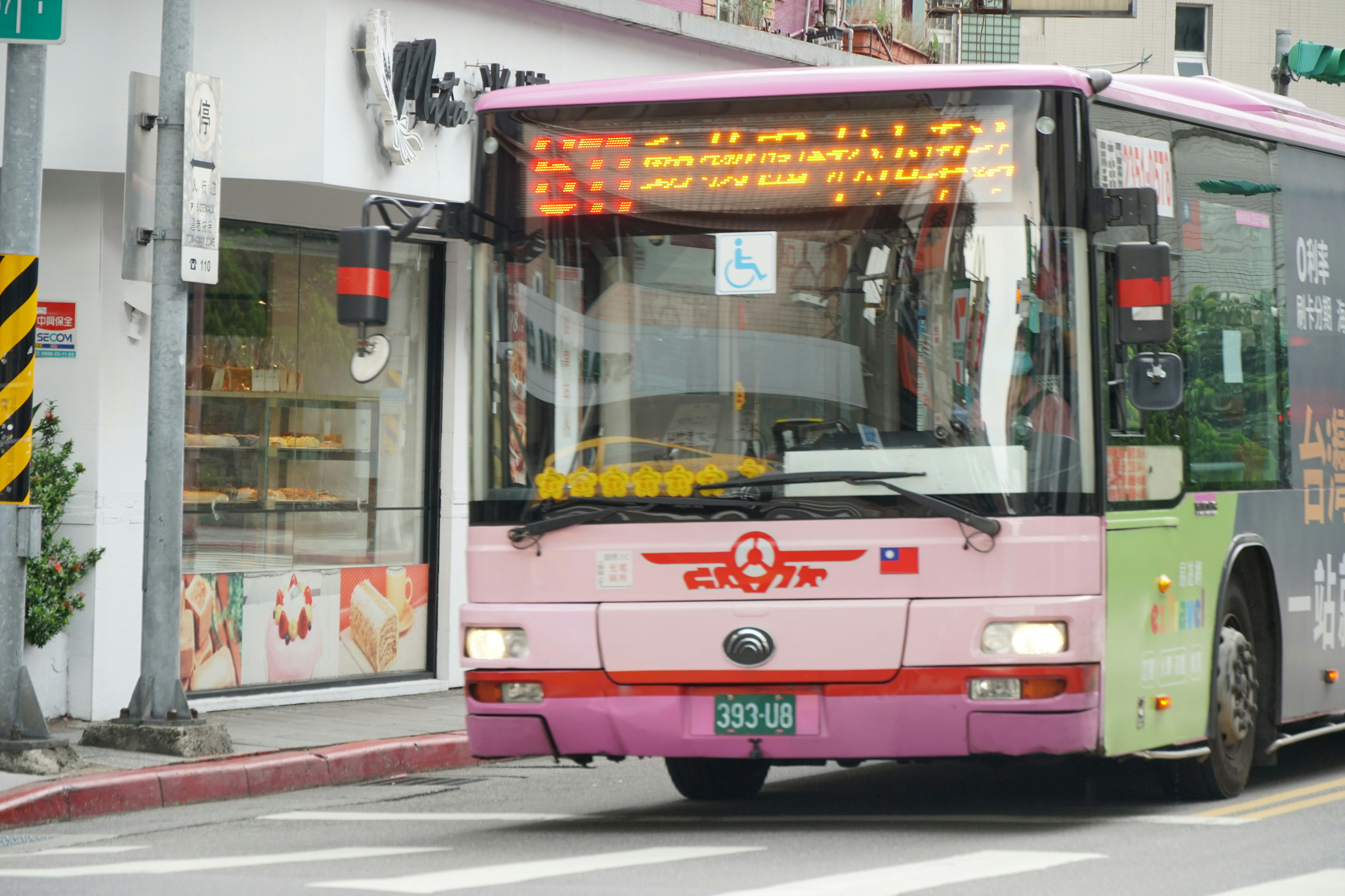 A pink city bus navigates a bustling street, showcasing digital route information and vibrant decorations. The backdrop features a bakery and urban architecture.