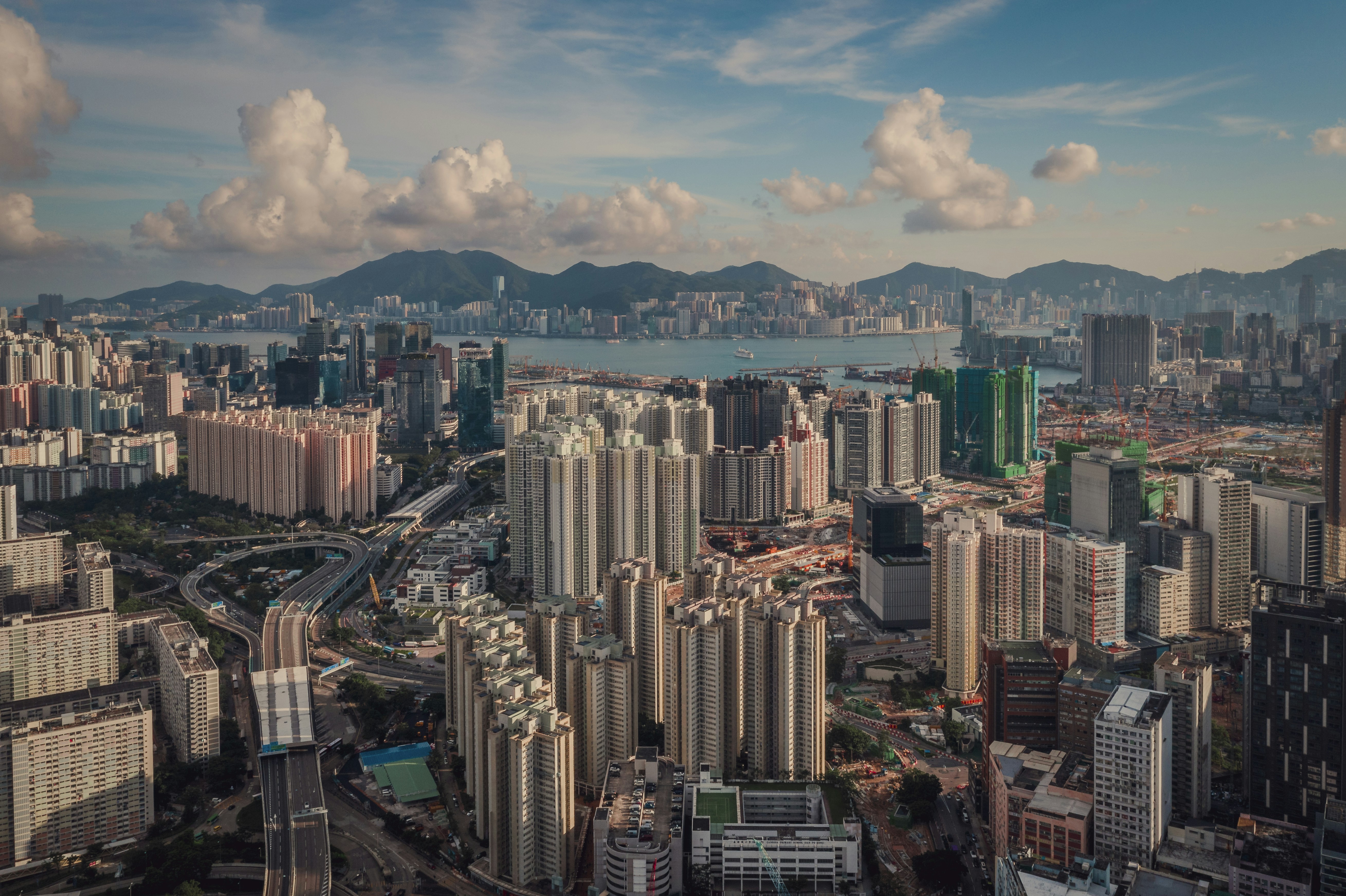 Aerial view of a sprawling cityscape with high-rise buildings, nestled between mountains and a shimmering bay under a cloudy sky.