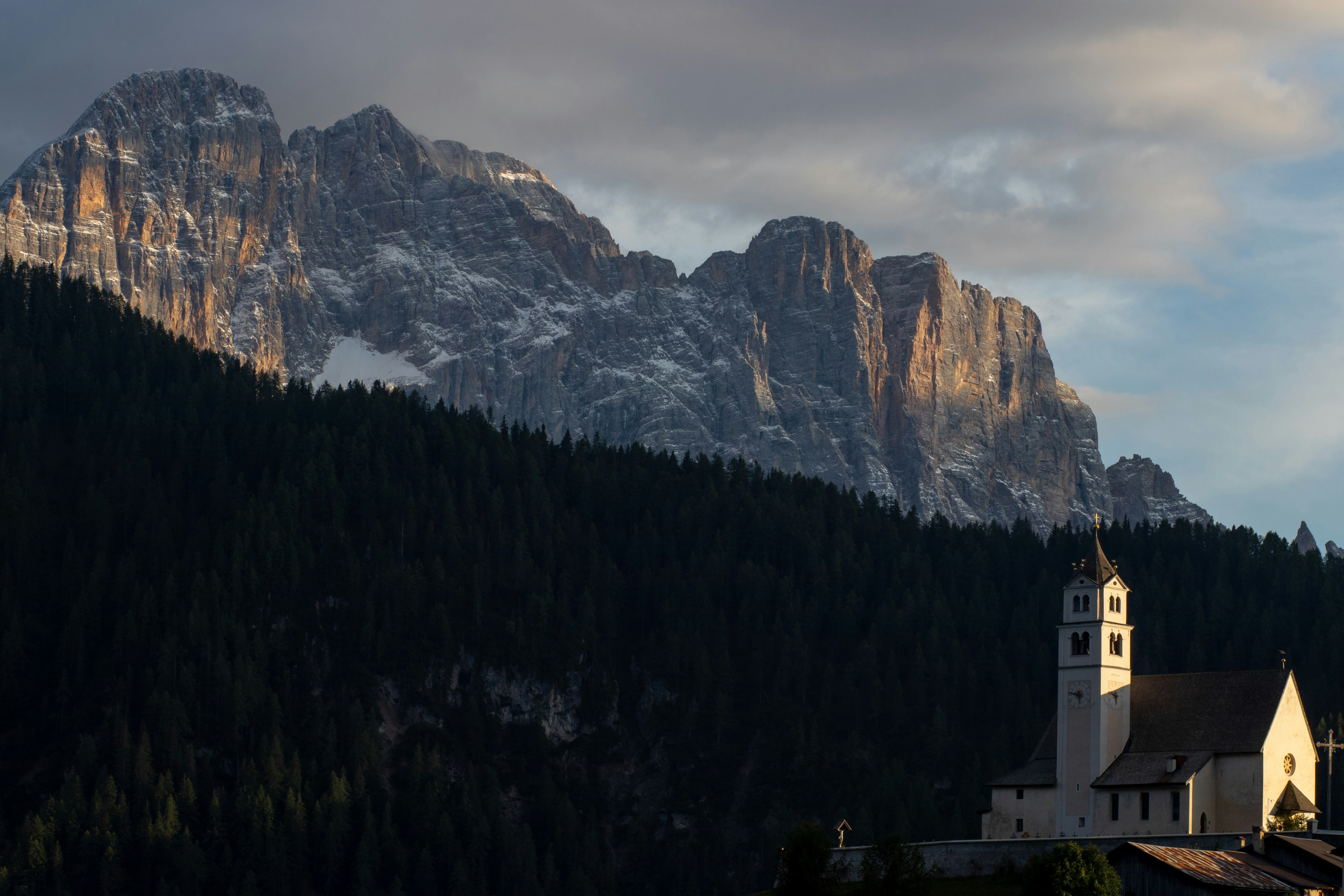 White church nestled against a forest with towering mountains in the background under a softly lit sky.
