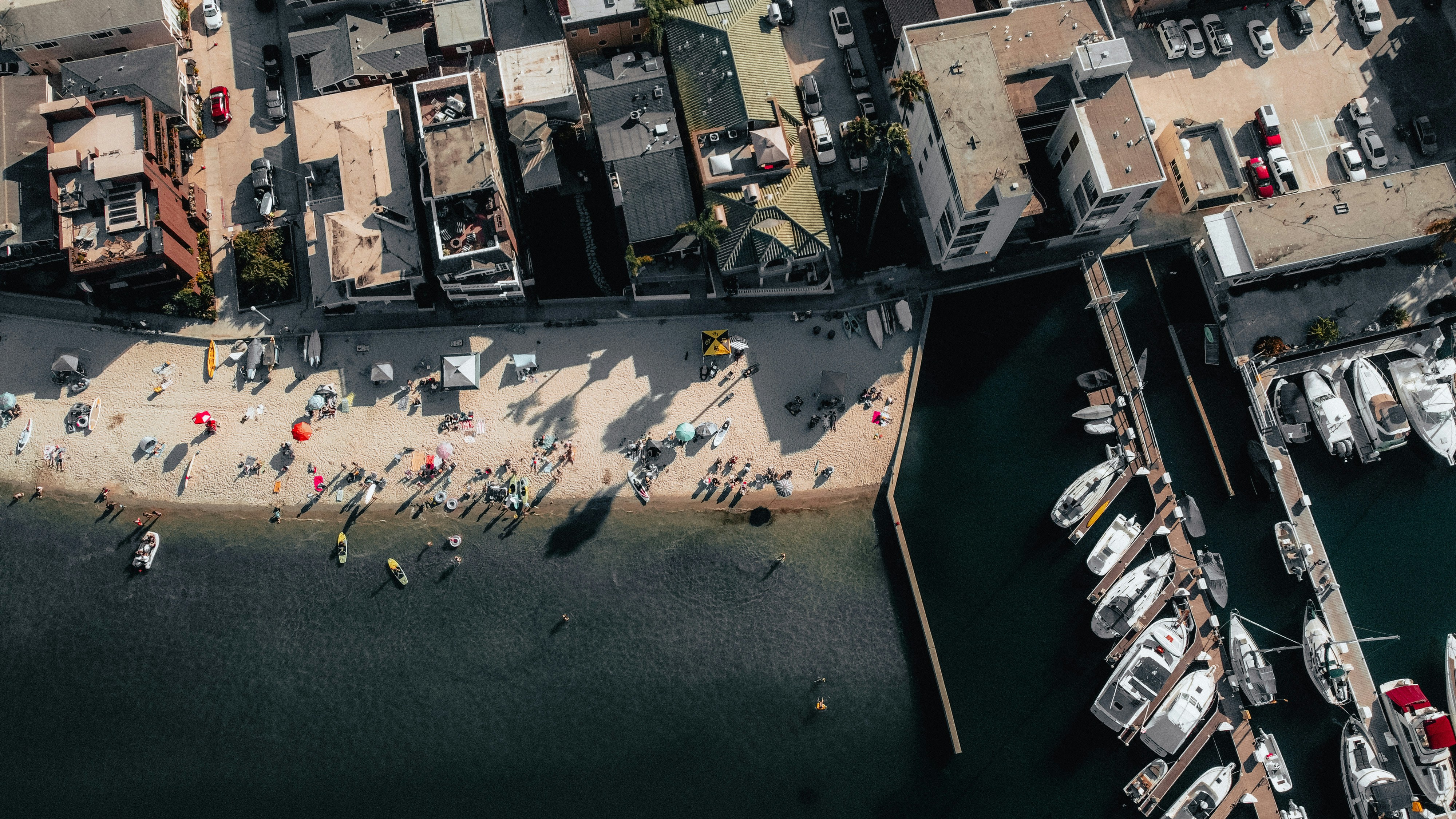 Aerial view of a bustling beach beside urban architecture and a marina with boats.