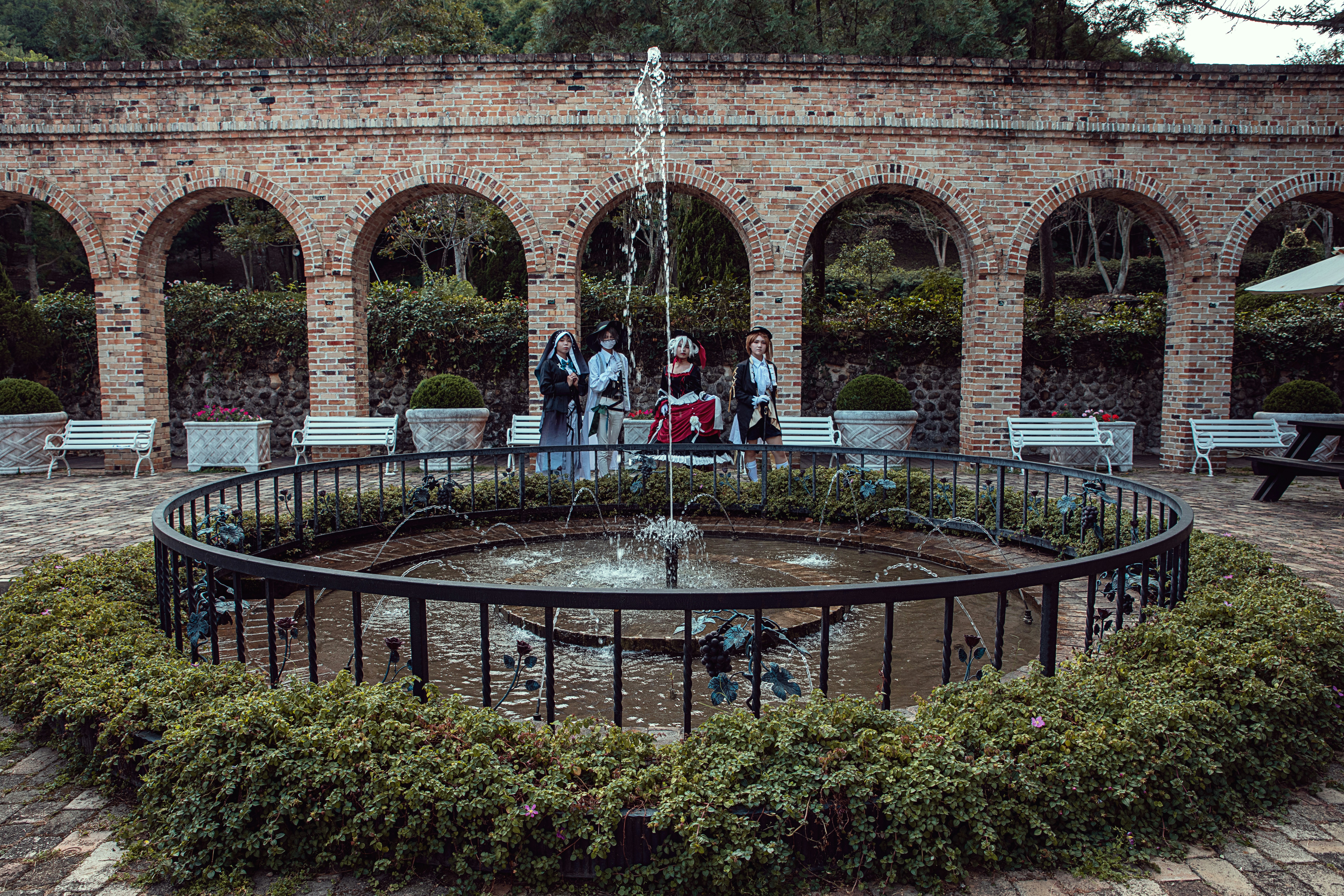 A group of friends enjoying a moment by a circular fountain surrounded by lush greenery and brick arches. The scene captures a tranquil atmosphere in a garden setting.