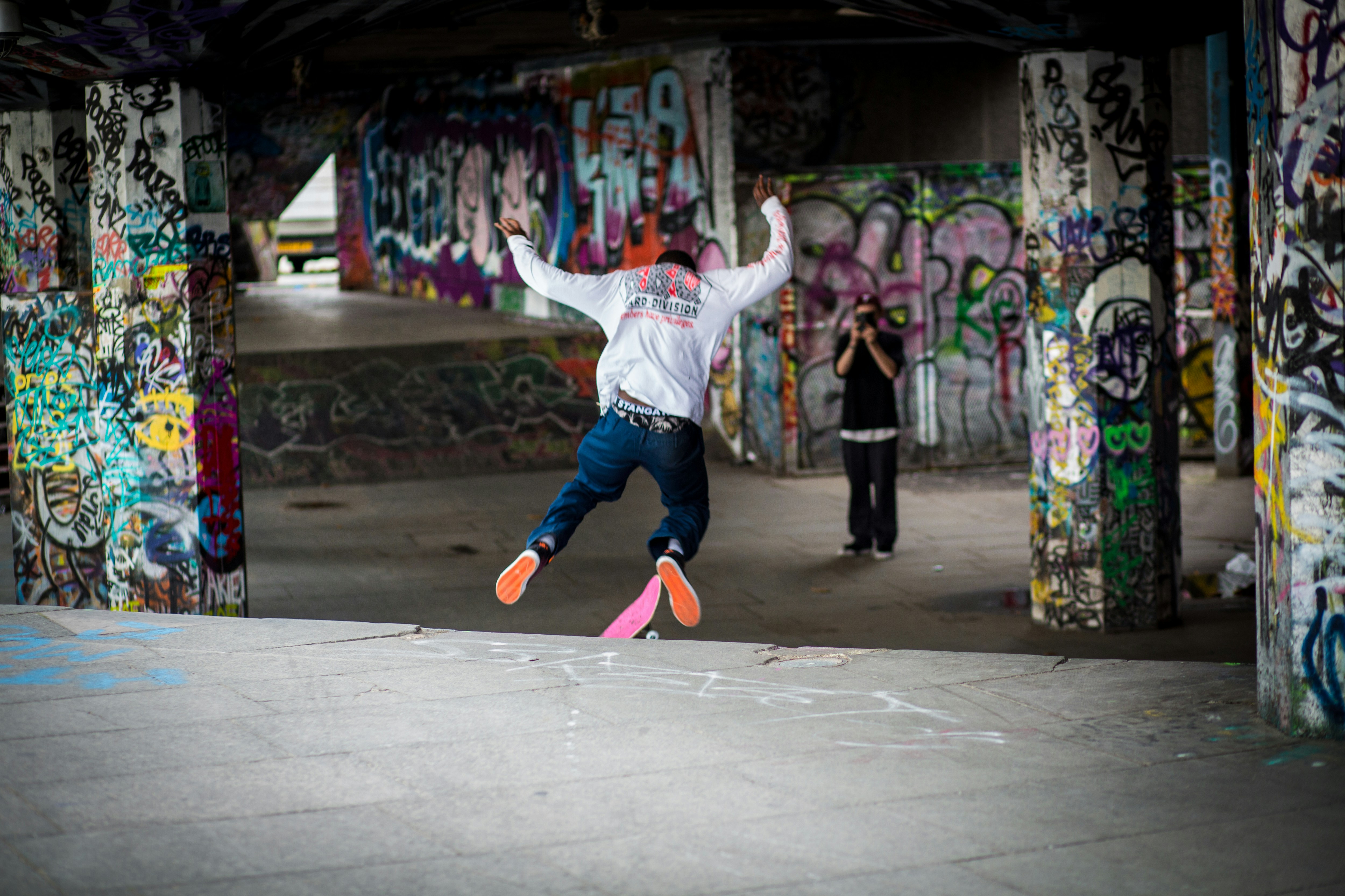 Skateboarder performing an airborne trick under a graffiti-covered overpass, with a spectator capturing the action in the background.