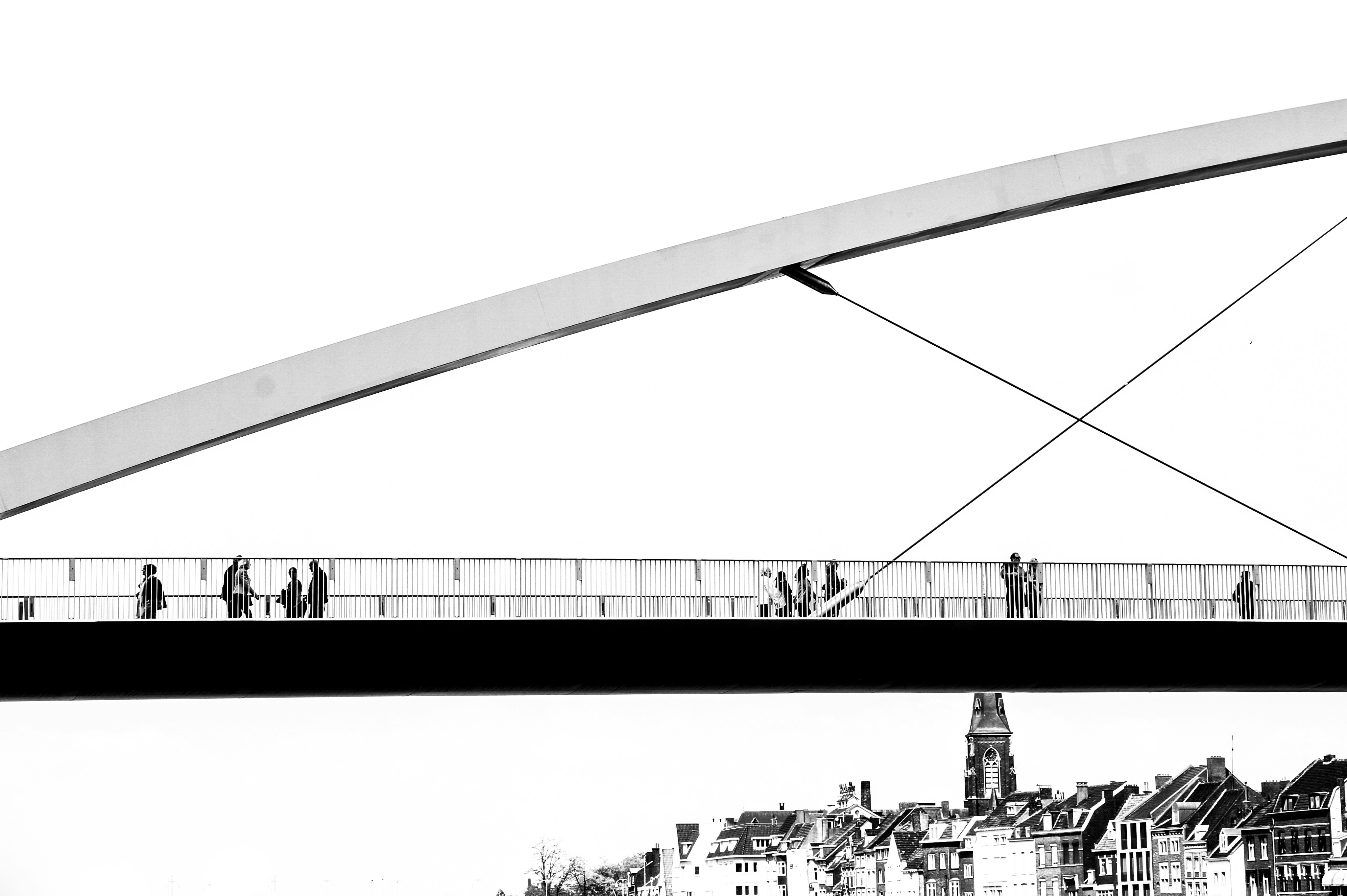 Black-and-white photograph of a curved pedestrian bridge with people strolling along its span. The lower city skyline provides contrast to the minimal upper scene.