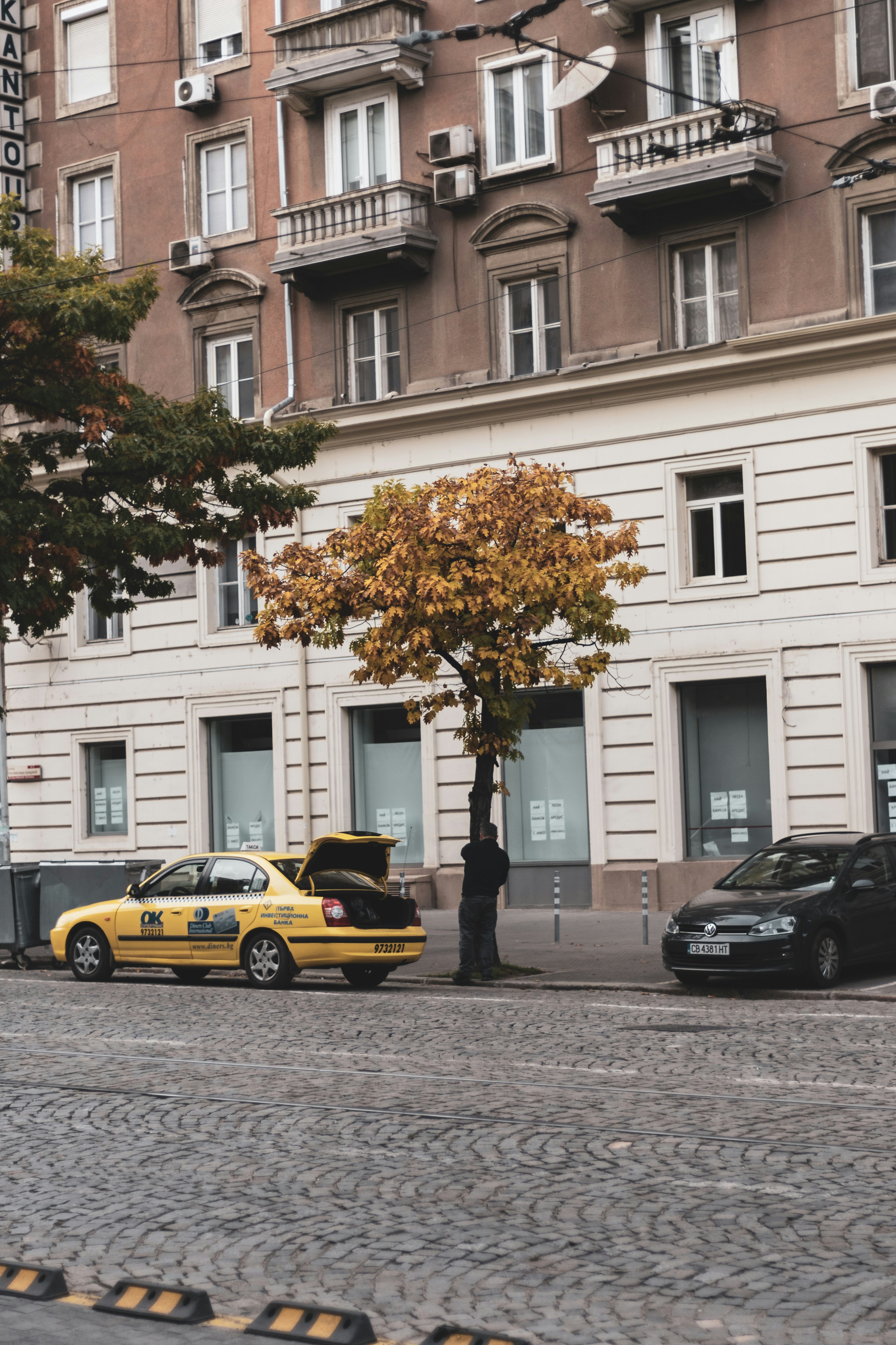 Yellow taxi parked beside a vibrant autumn tree on a cobblestone street, with a classic building in the background.