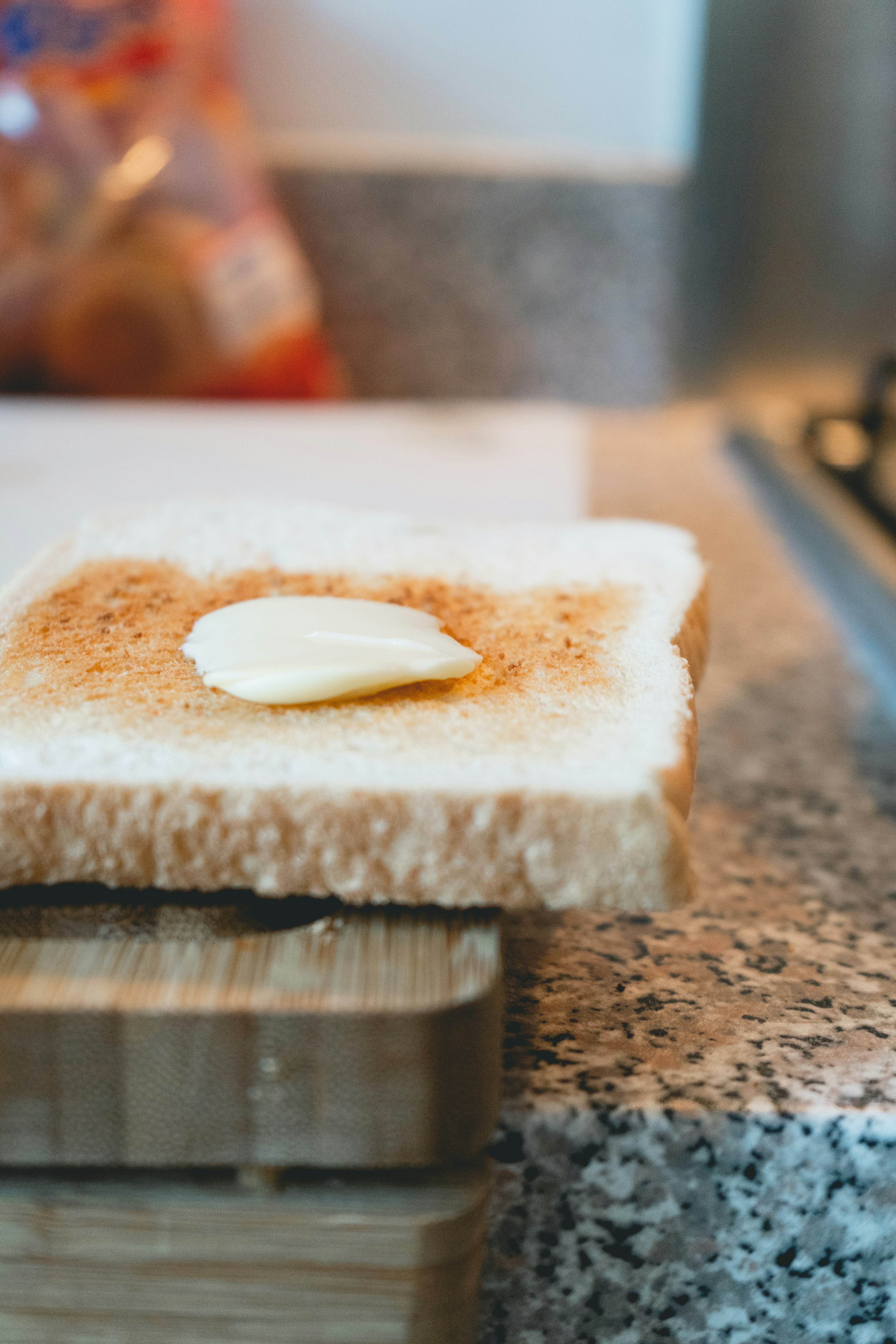 A slice of toasted bread topped with a dollop of butter, resting on a wooden cutting board against a blurred kitchen background.