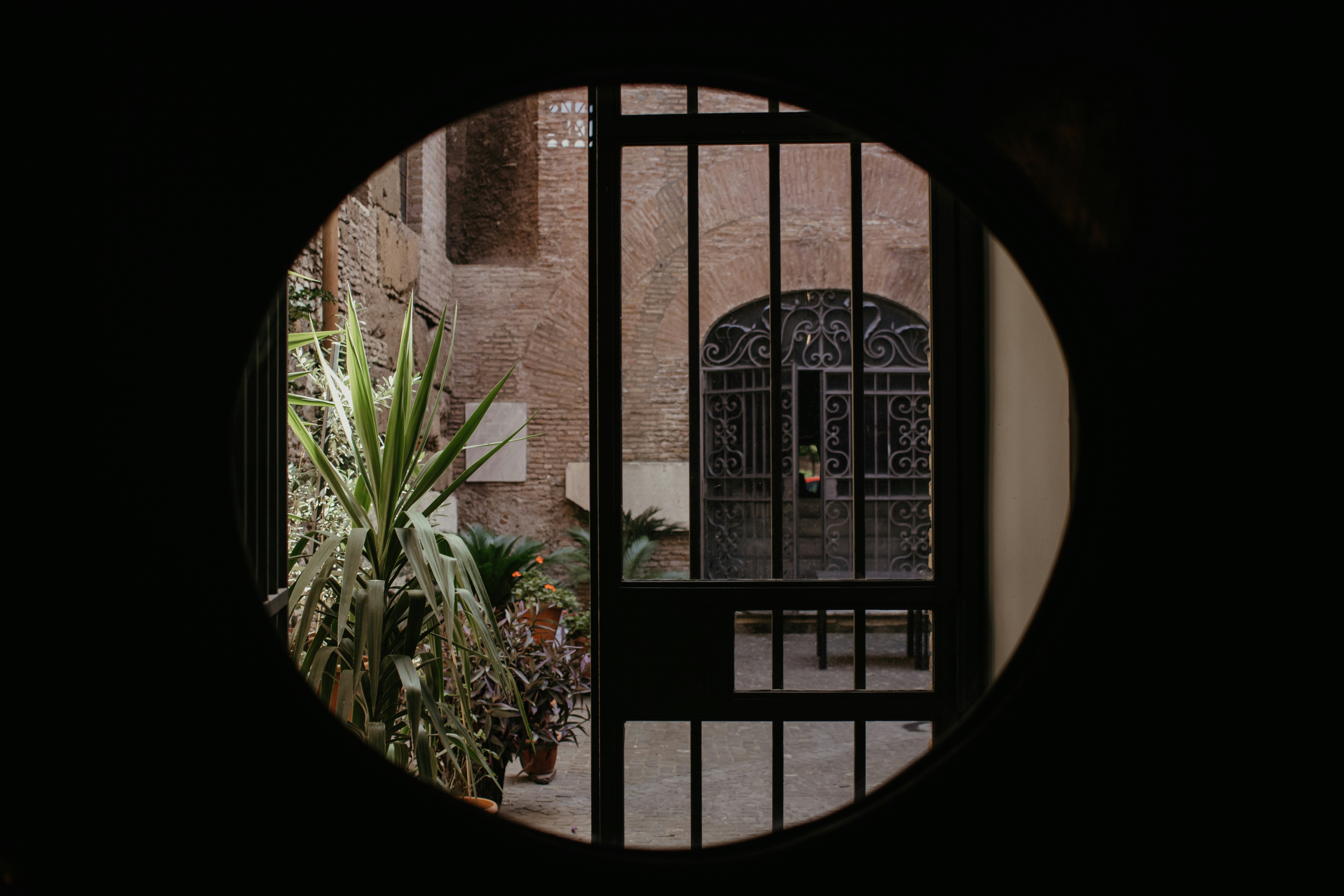 View through a circular opening showcasing a lush courtyard with greenery and an ornate gate. The composition highlights the contrast between indoor and outdoor spaces.