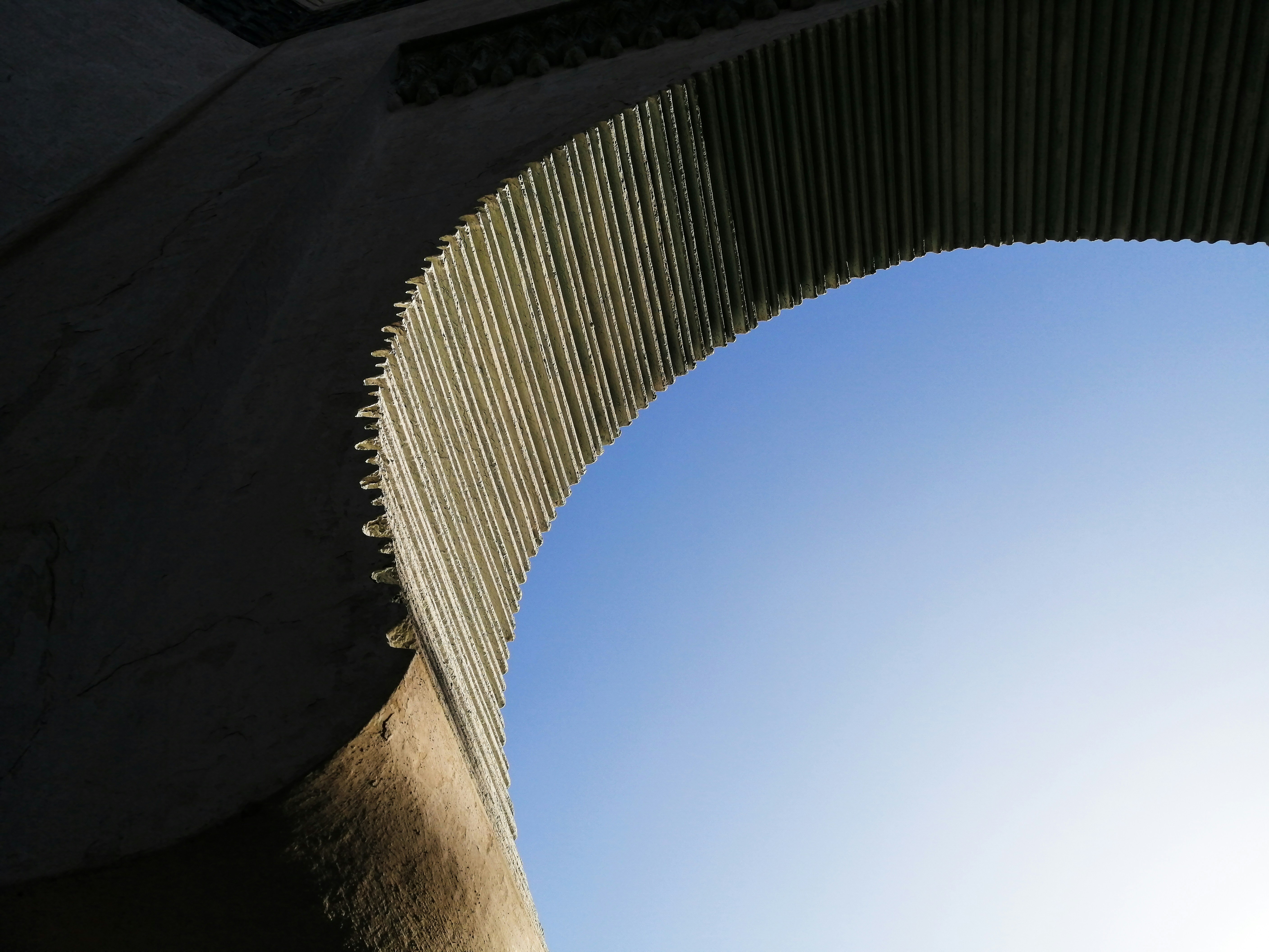 Curved architectural structure framing a clear blue sky, showcasing intricate lines and textures. The play of light enhances the design's depth.