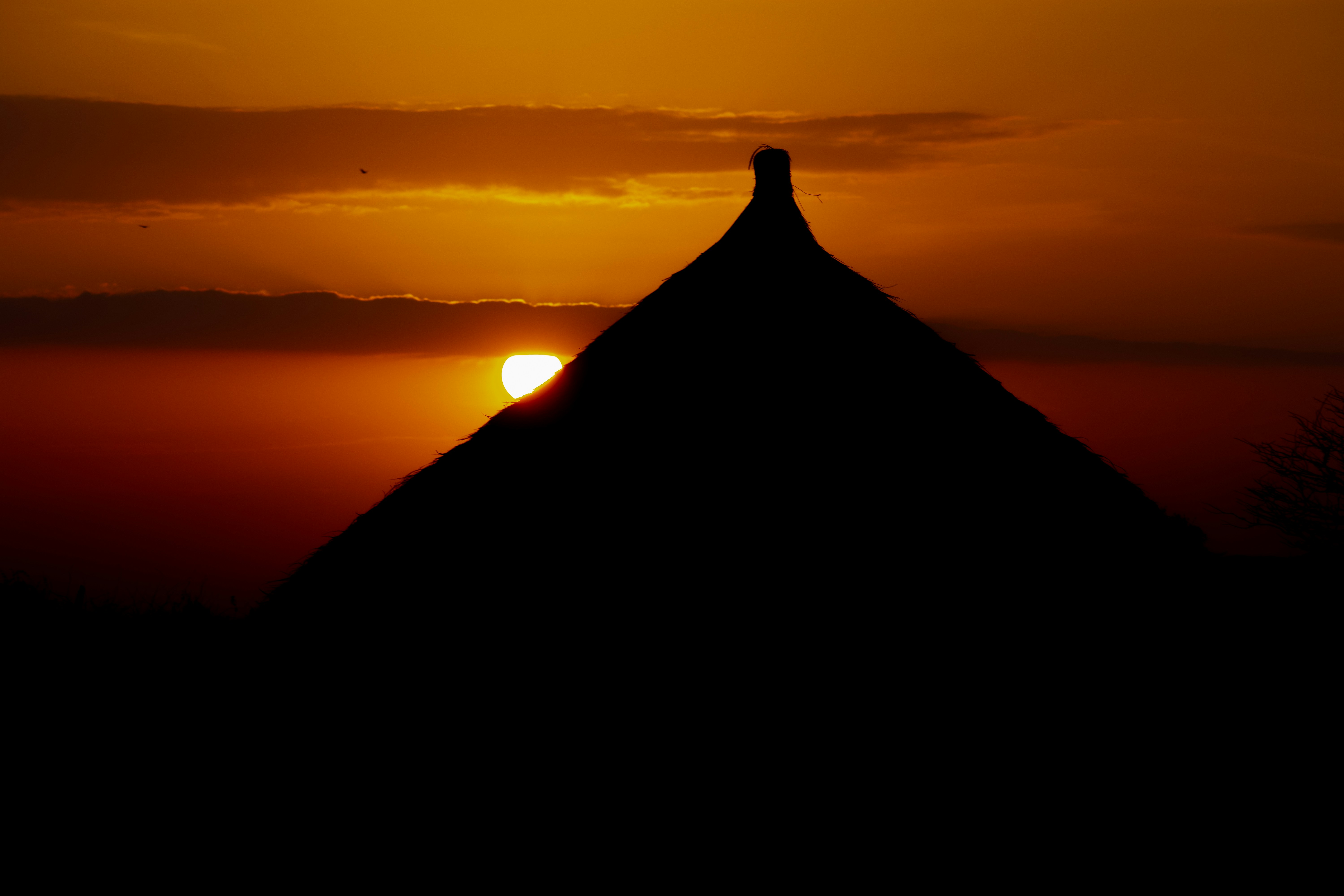 Traditional thatched hut silhouetted against a vibrant sunset, with the sun partially hidden behind the horizon. 