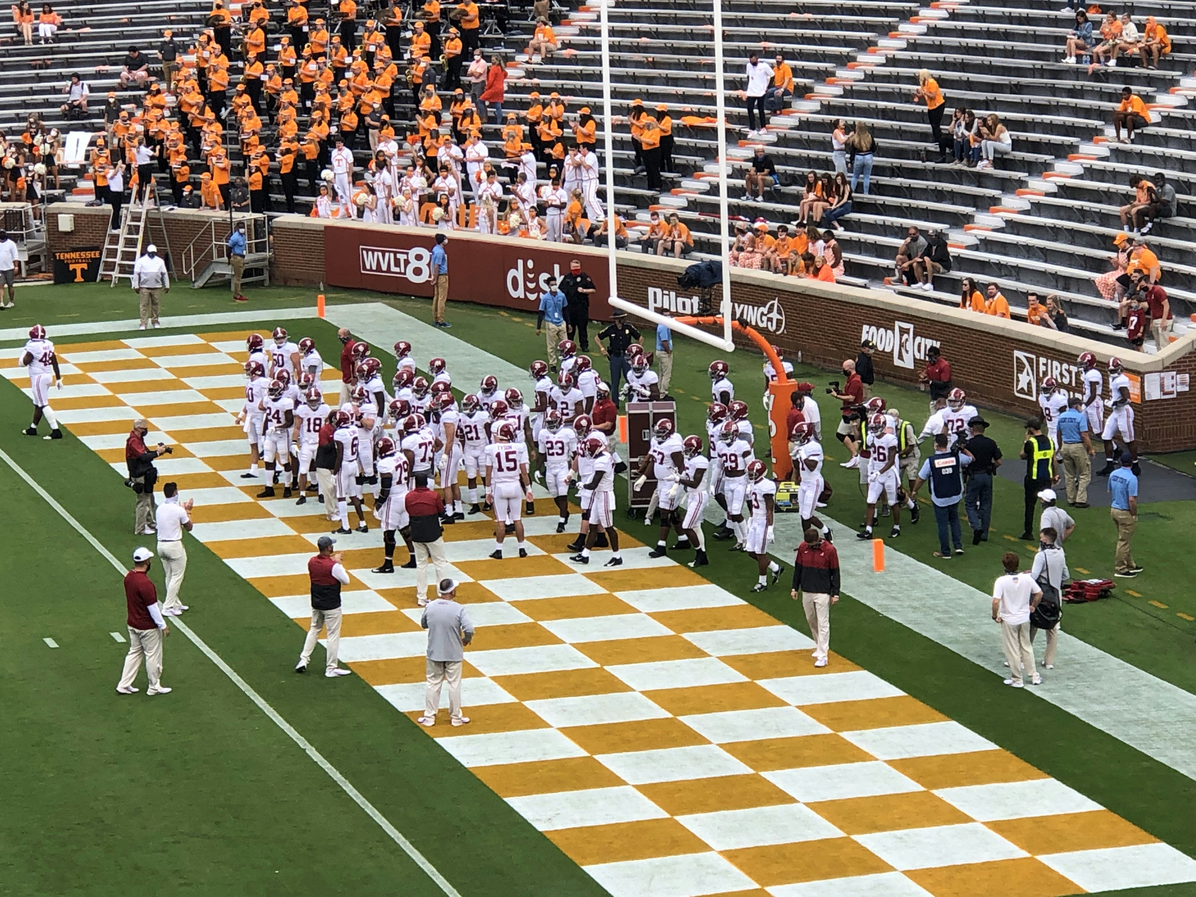 Football team huddles in the end zone of a stadium before a game, with fans in orange shirts filling the stands.