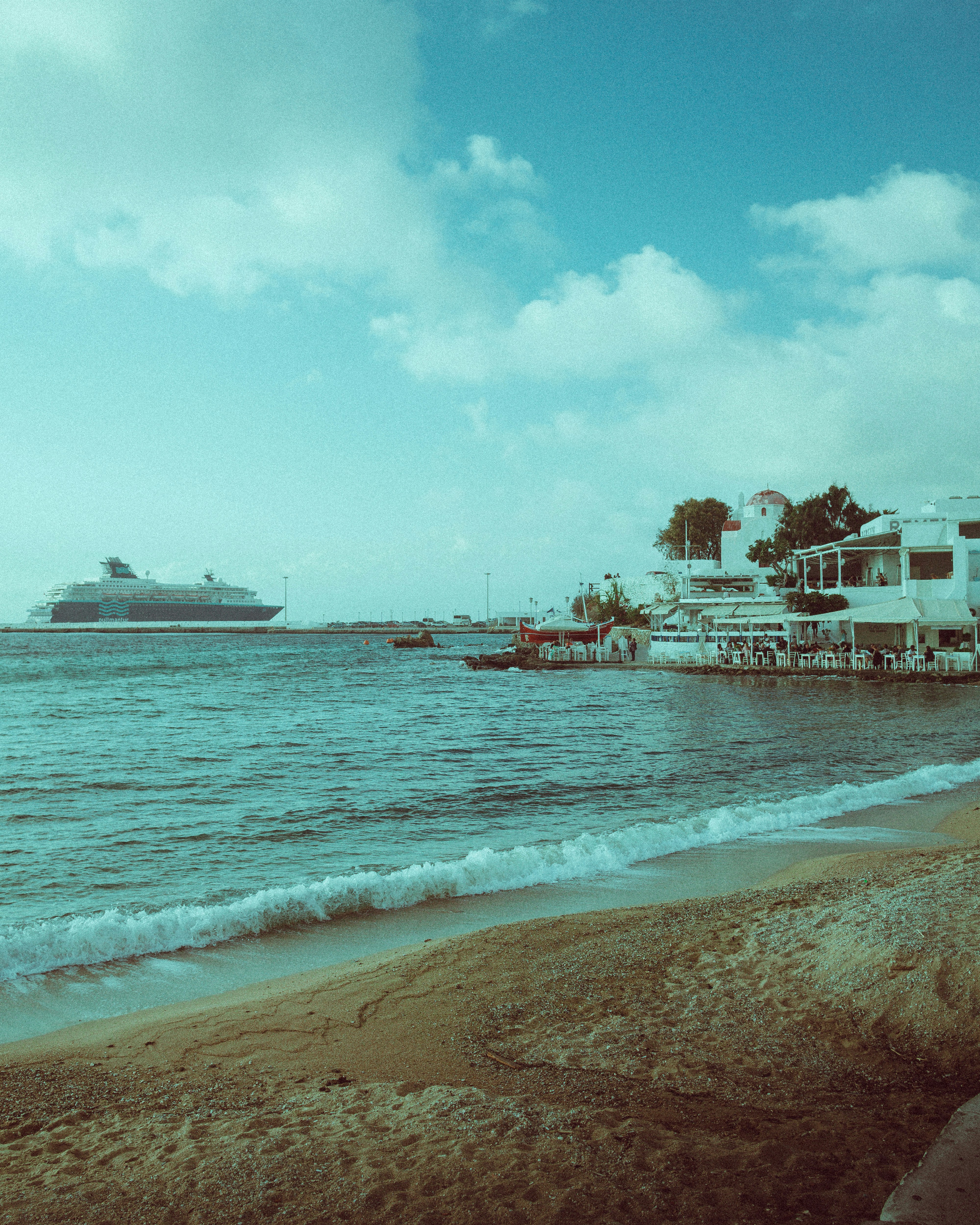 Cruise ship anchored in a tranquil harbor with sandy shores and gentle waves, framed by a picturesque waterfront establishment.