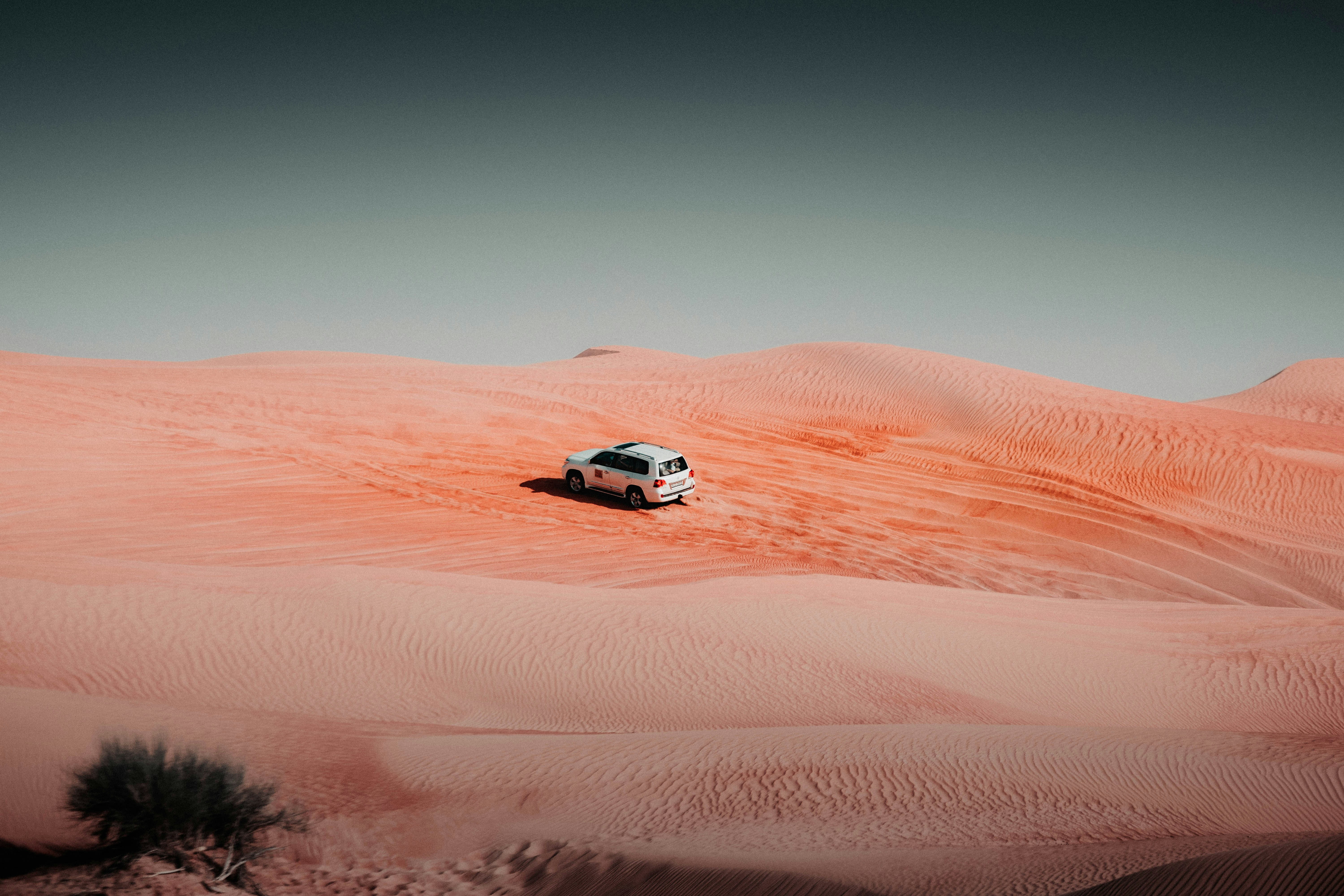A lone vehicle navigates the vast expanse of a sunlit desert, surrounded by undulating sand dunes. The scene captures the essence of adventure and isolation.