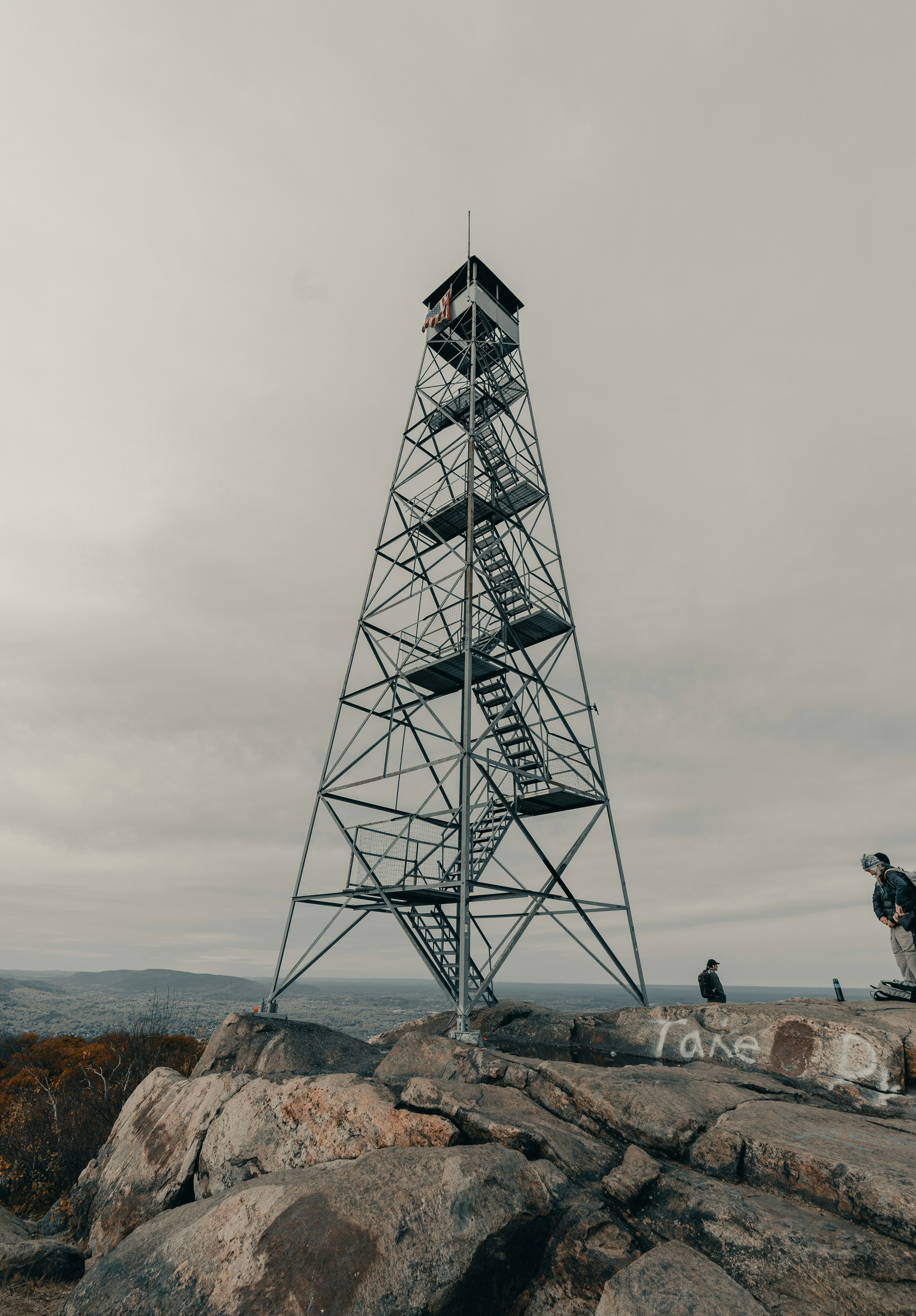 Steel fire tower standing tall on rocky terrain, surrounded by expansive views of the landscape. A few hikers explore the area below.