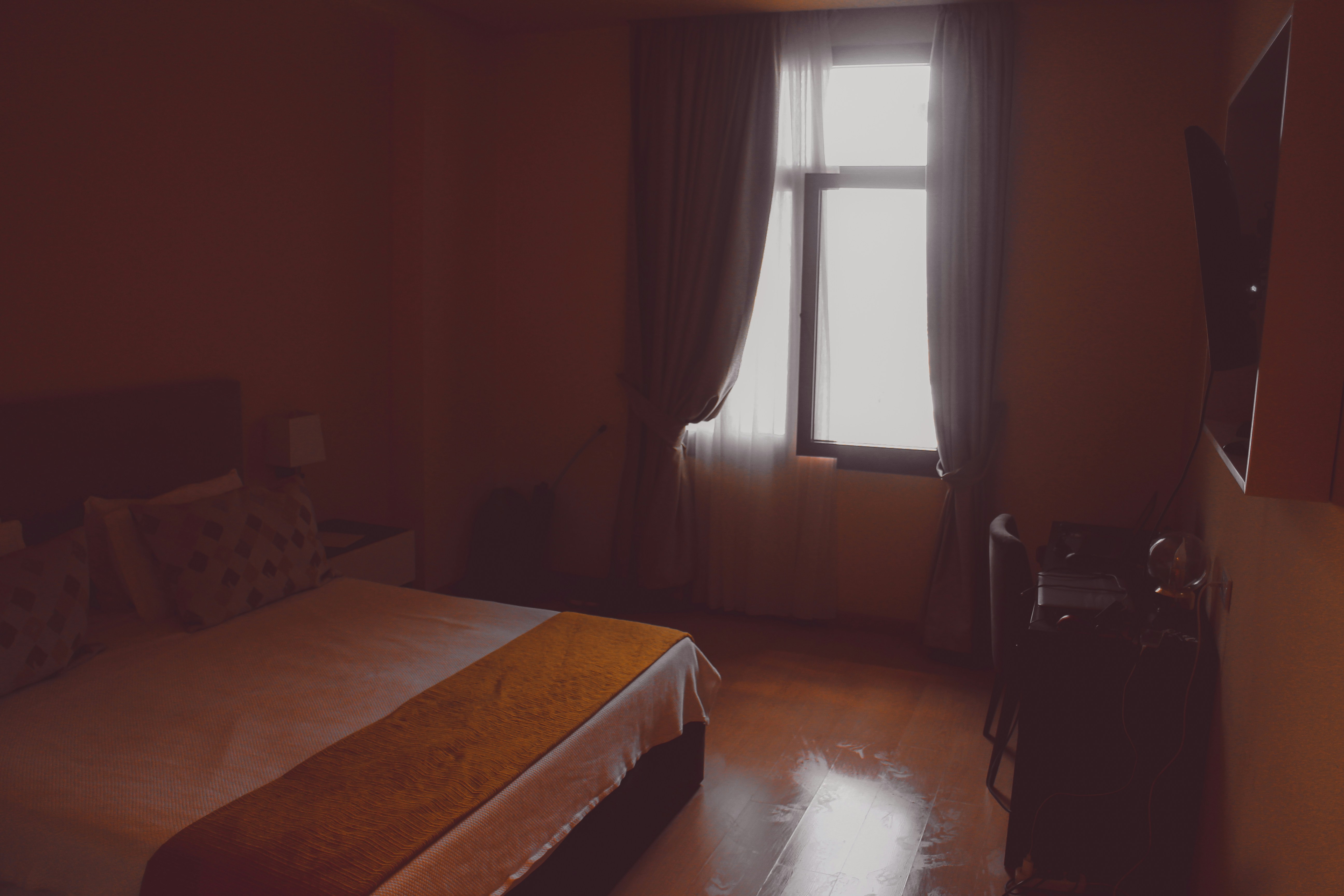Cozy hotel room with a neatly made bed and soft lighting, featuring a large window draped with curtains. A small desk and chair are visible in the corner.