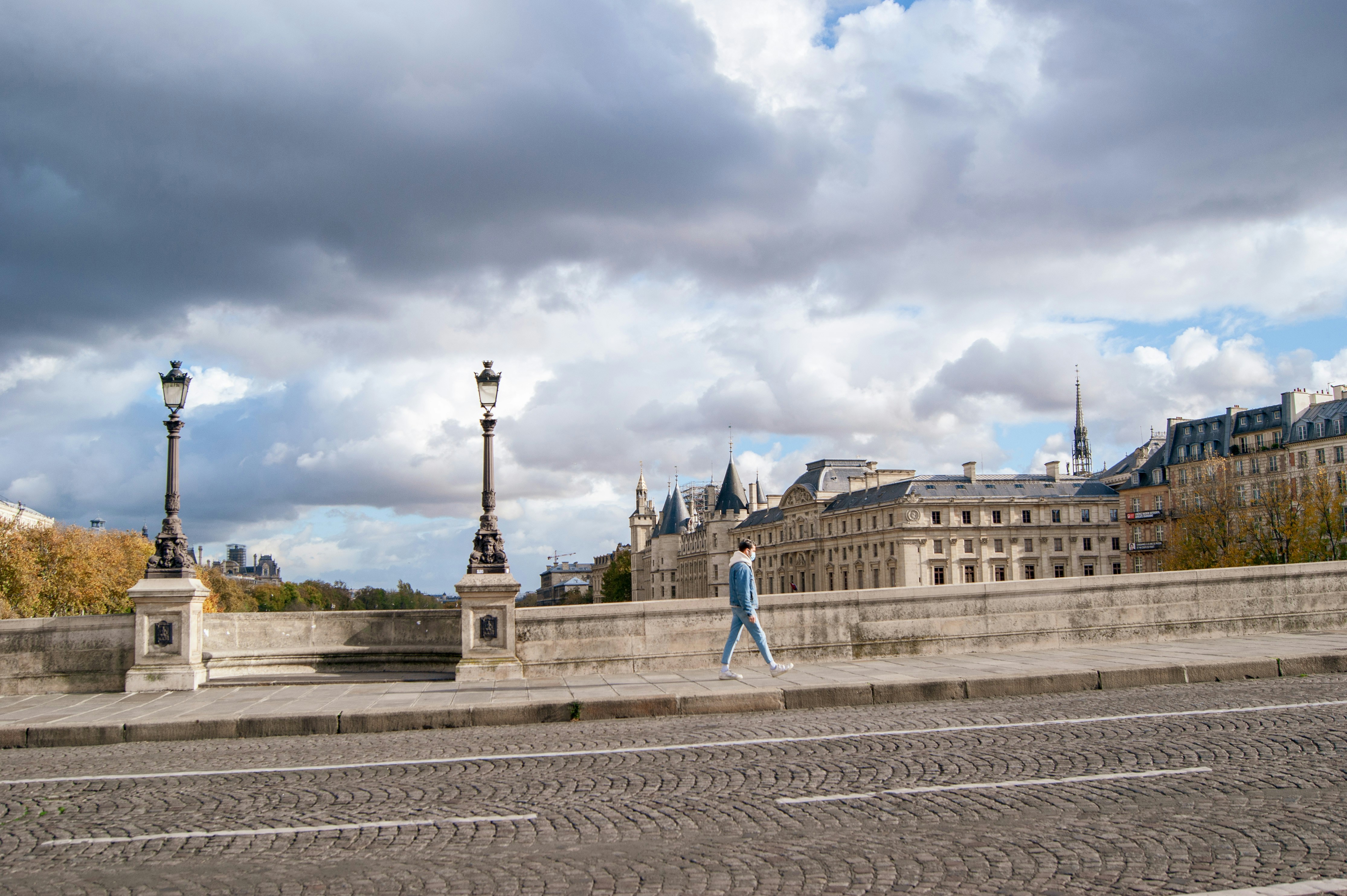 Blue-jacket pedestrian walks along a cobblestone Parisian bridge, flanked by classic lamp posts with historic buildings in the distance under a cloudy sky.