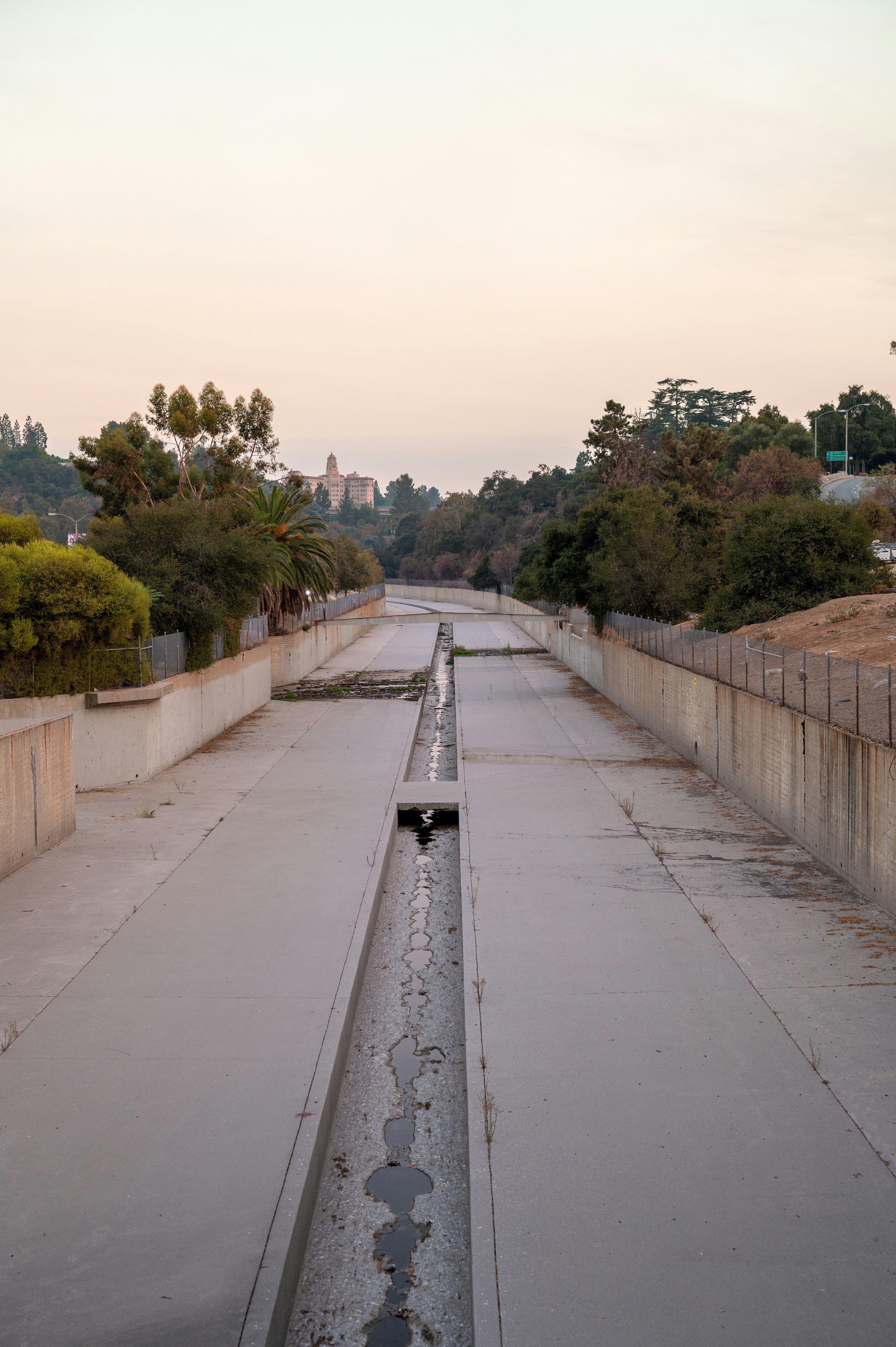 Concrete waterway lined with vegetation, leading towards distant buildings under a soft evening sky.