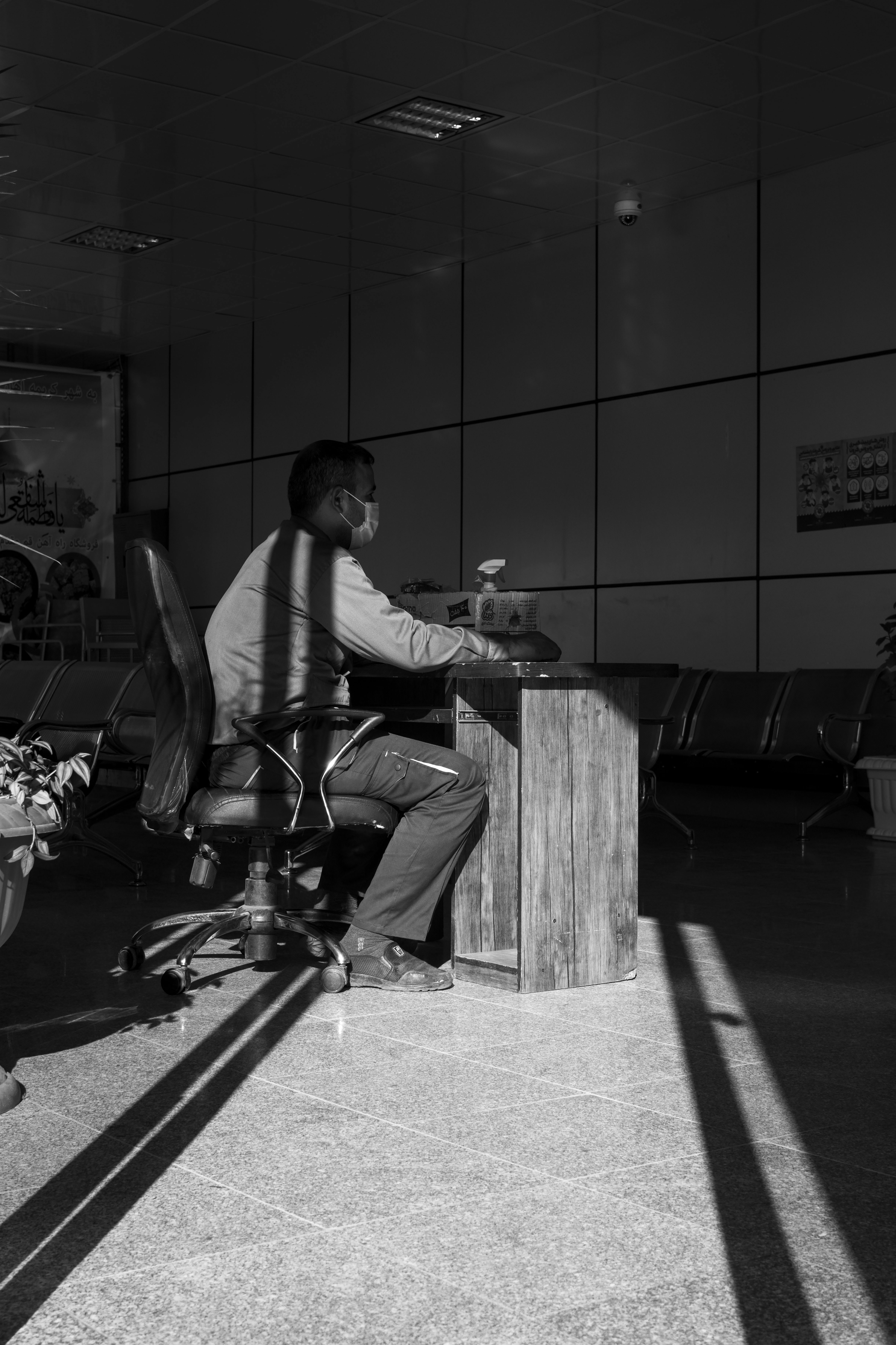Shadowed Silence: A Masked Clerk at a Wooden Desk Under Striped Light