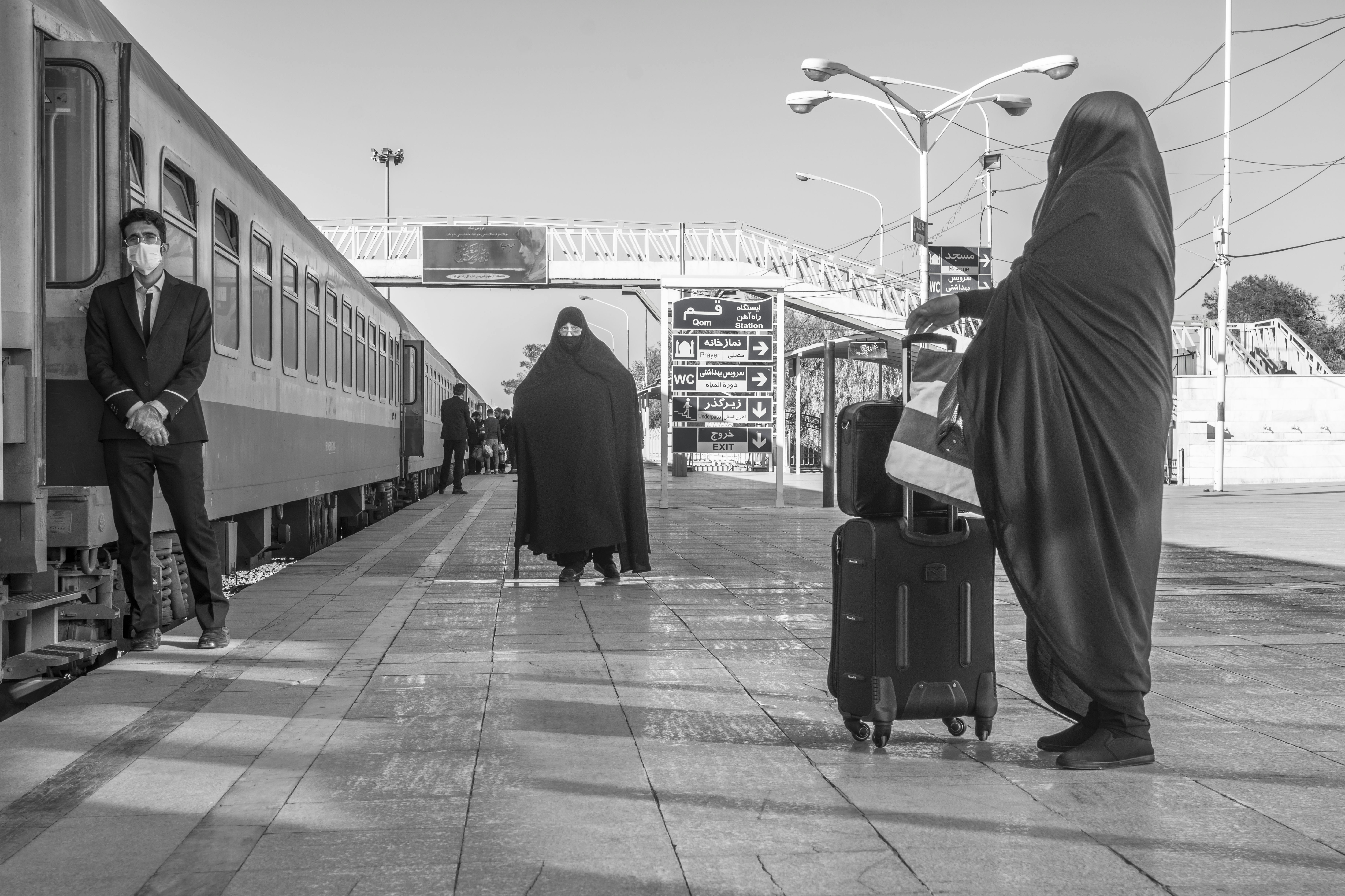 Two women in traditional attire await their train at a bustling station, while a man in formal wear stands nearby, creating a contrast of styles and stories.