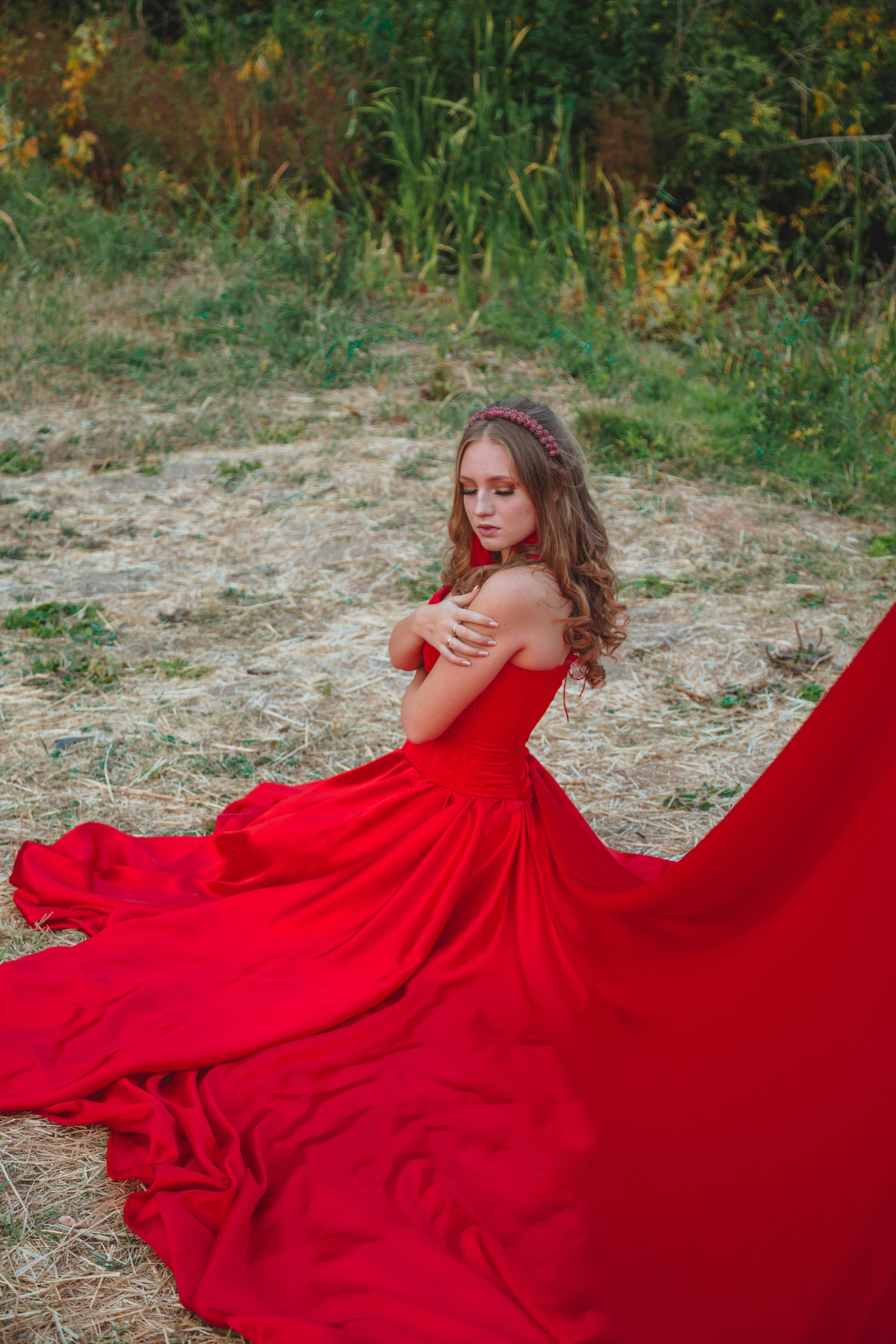 A young woman in a flowing red gown poses gracefully amidst a natural backdrop, embodying elegance and serenity.