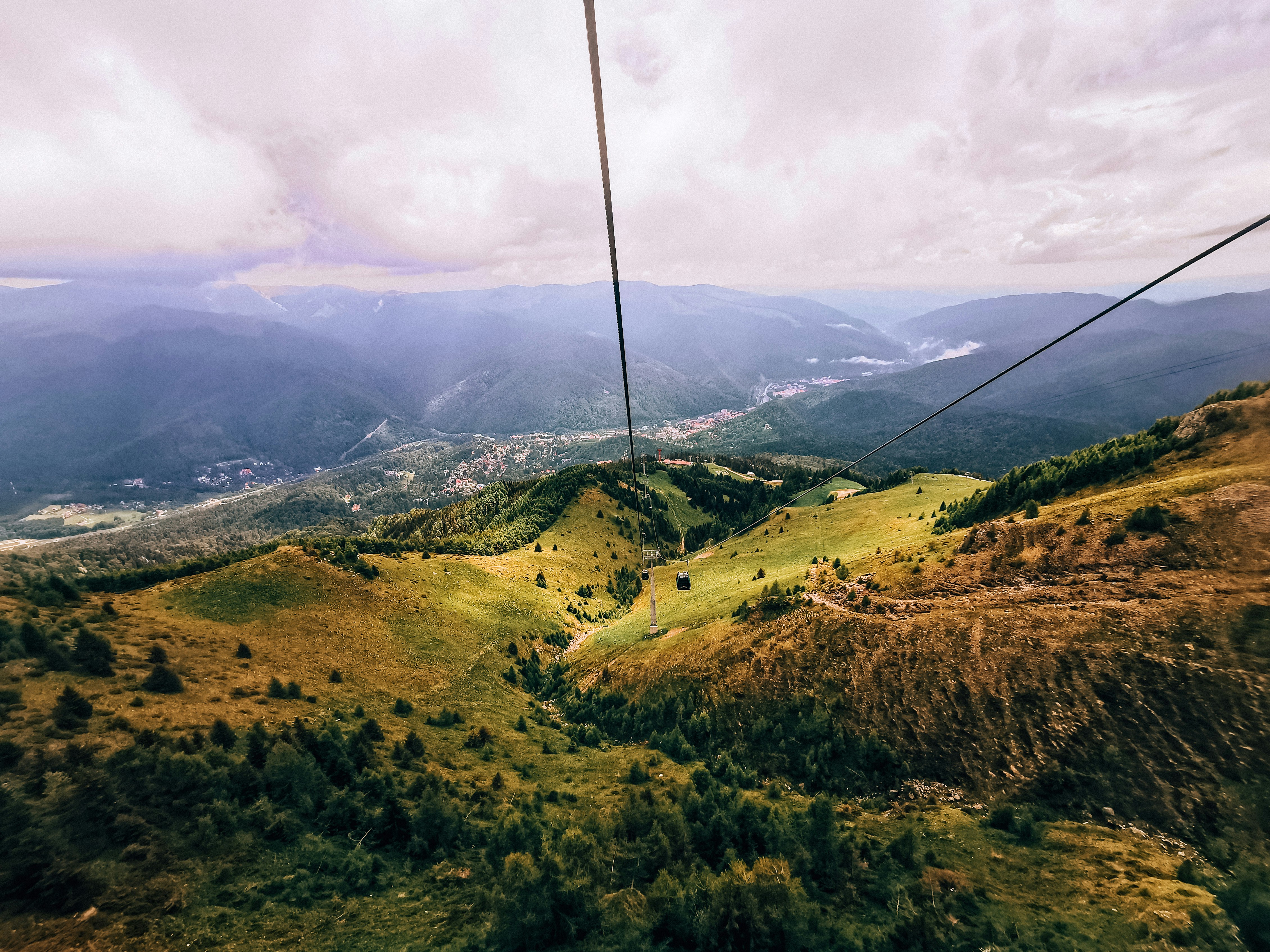 Expansive view of rolling green hills and valleys beneath a cloudy sky, intersected by cable lines.