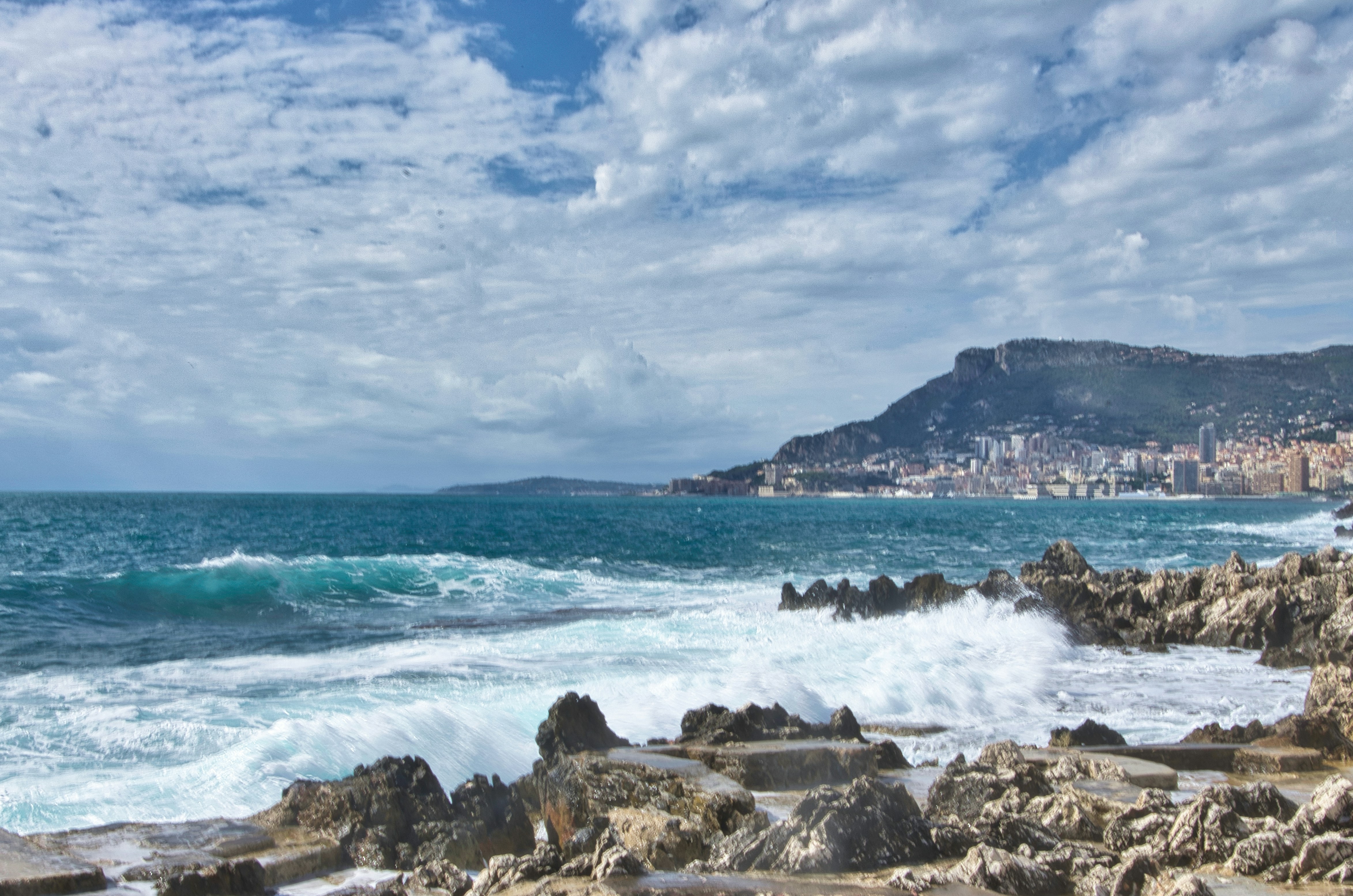 Rugged coastline of Cap Martin with waves crashing against rocks under a partly cloudy sky.