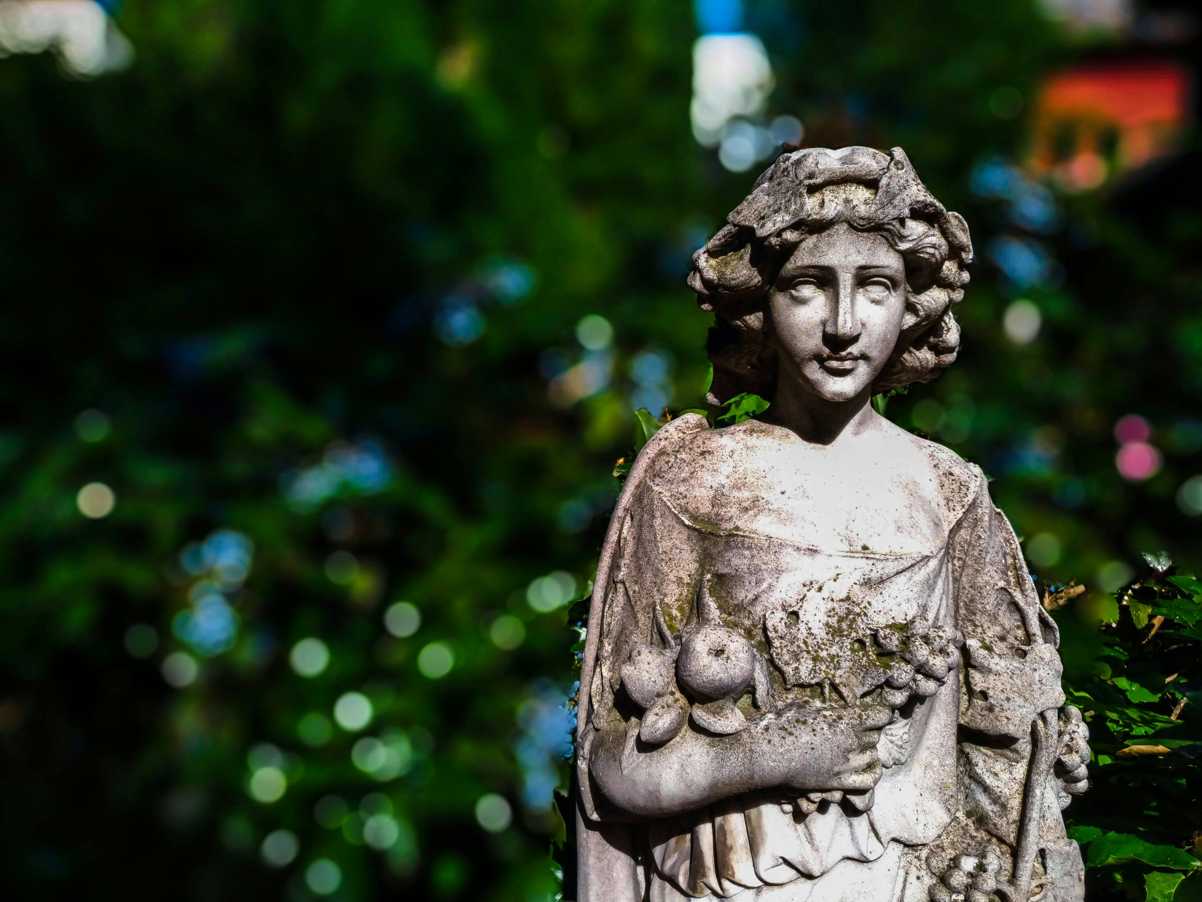 A weathered statue of a woman holding fruits, surrounded by a blurred, lush backdrop, evoking a sense of tranquility and history.