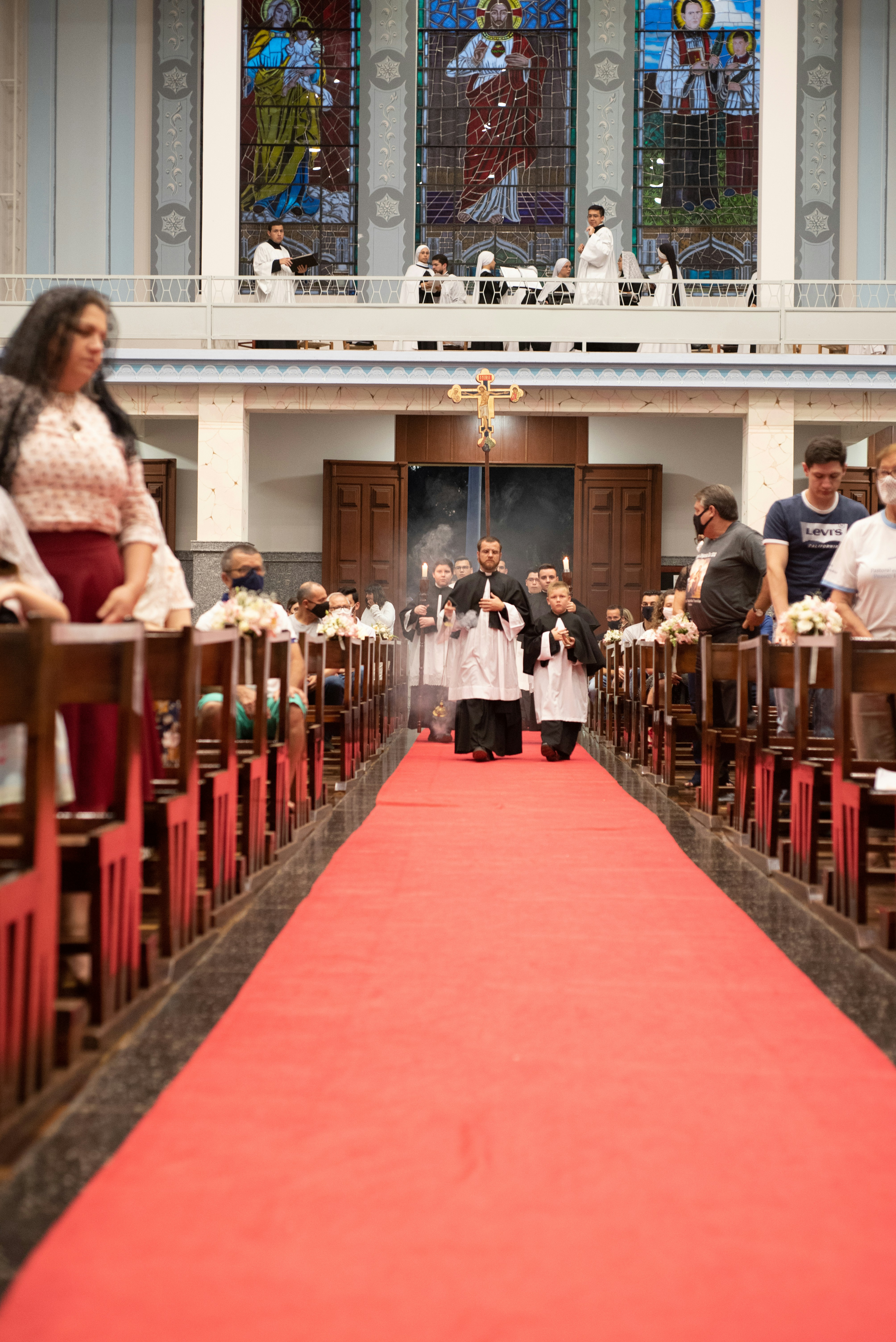 Clergy leading a procession down a red carpet in a church, with stained glass windows in the background.